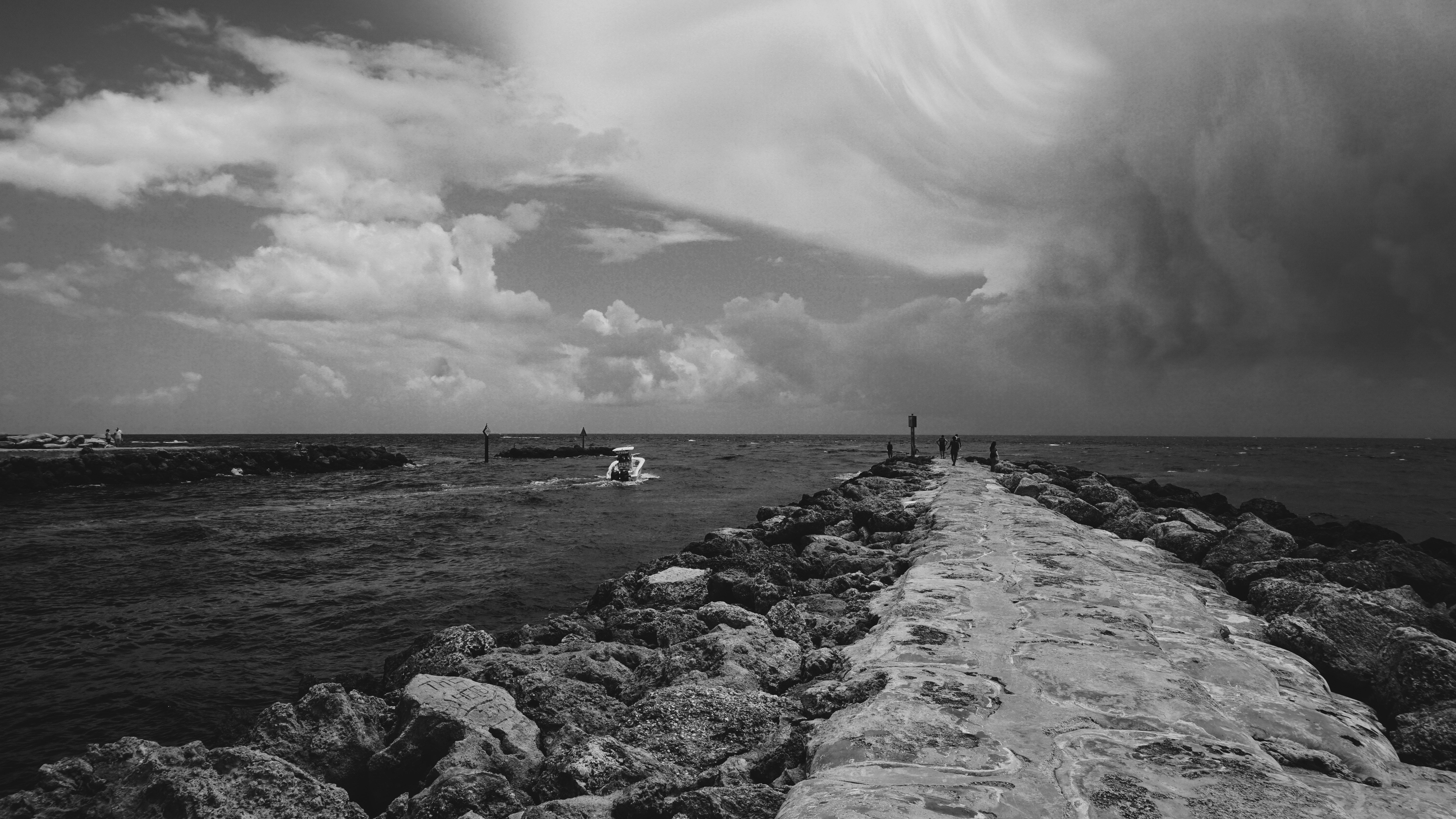 Dramatic black and white seascape with rocky pier leading into the ocean under a cloudy sky.