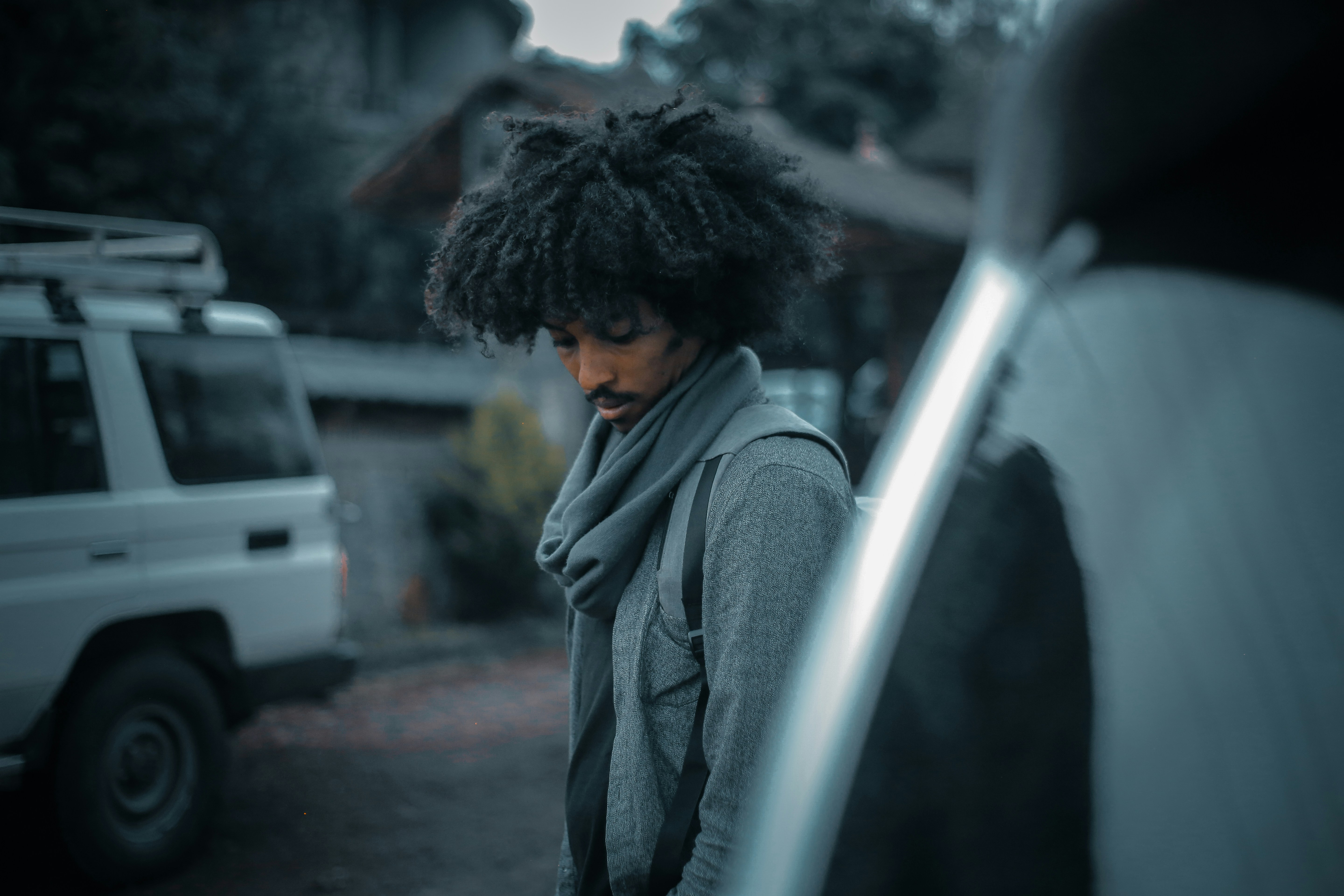 A man with voluminous hair stands pensively beside a vehicle, enveloped in a serene atmosphere that hints at introspection.
