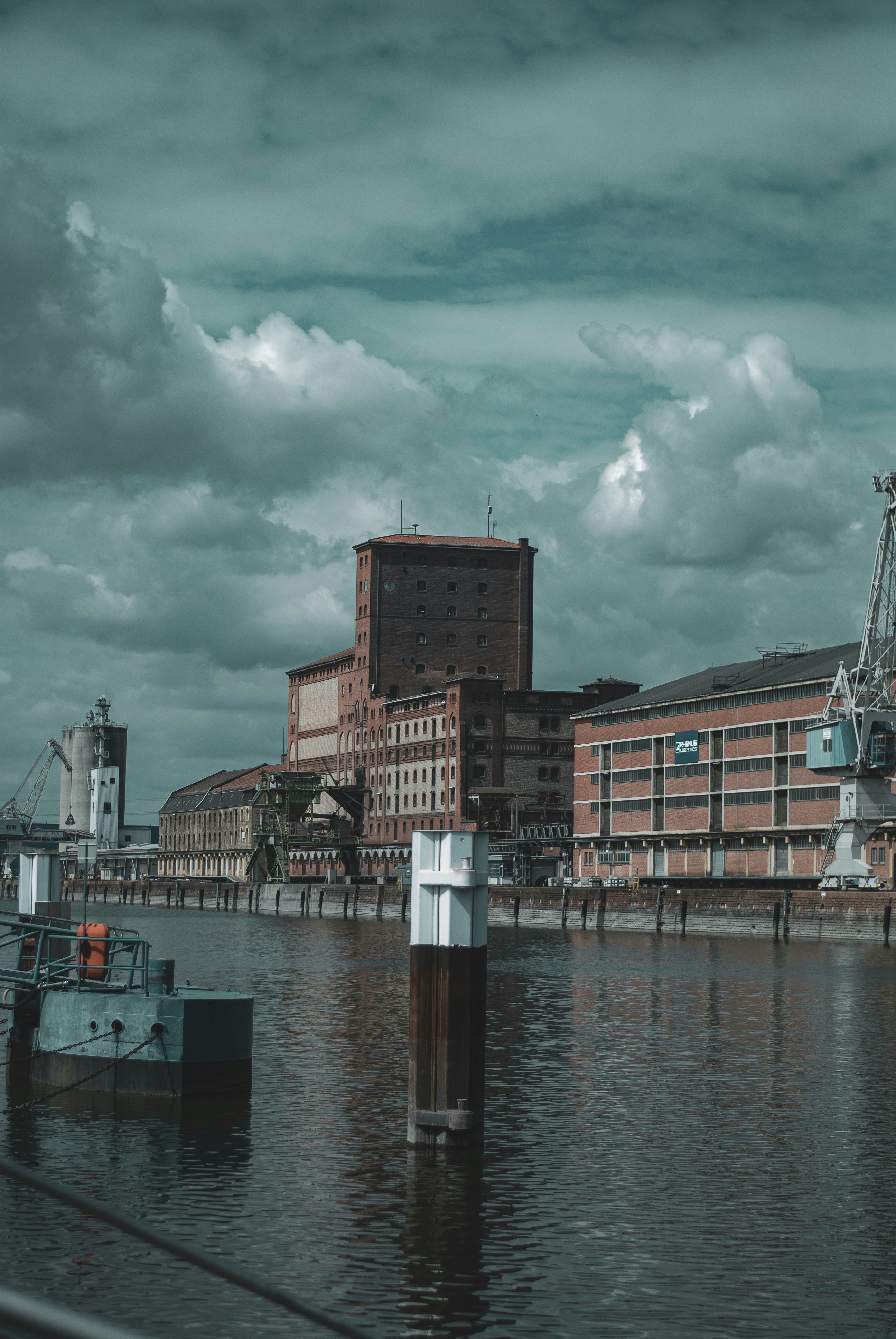 Historic warehouses and a crane reflected in calm waters under a moody sky, showcasing the remnants of a bustling port.