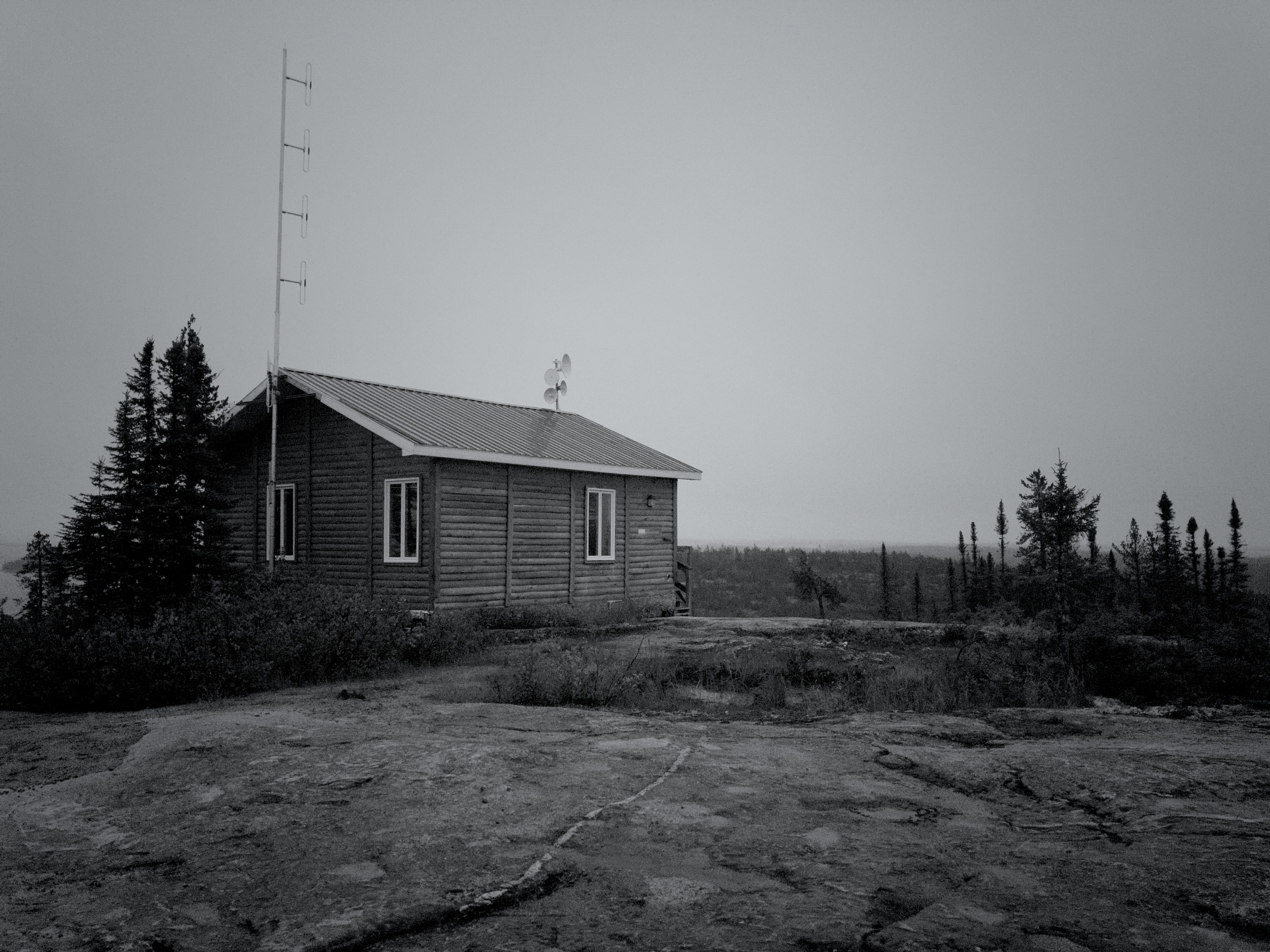 Weathered wooden cabin stands on rocky terrain, surrounded by dense pines under a gray sky. A communication tower rises beside it.