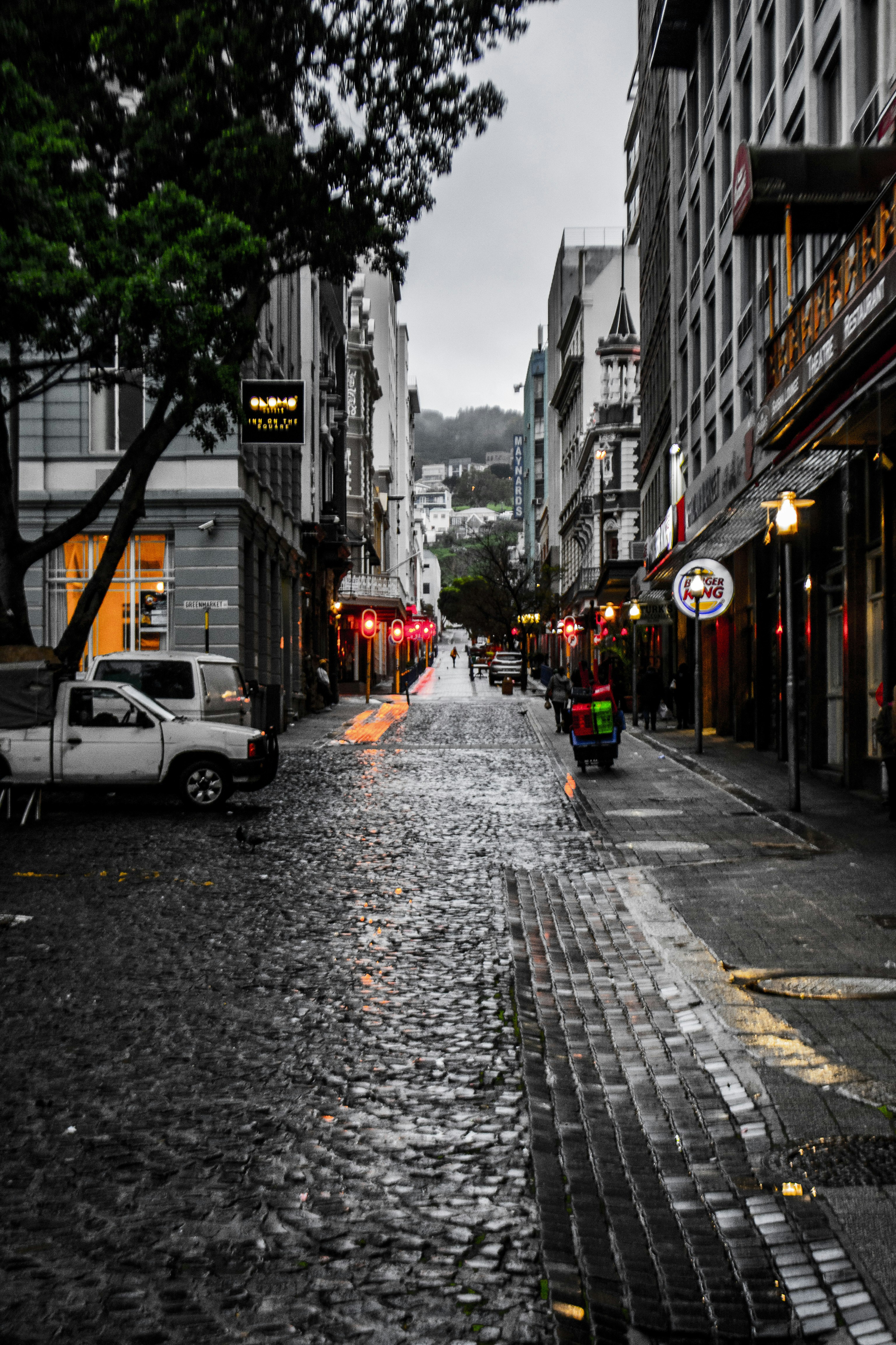 Cobblestone street glistening after rain, lined with buildings and illuminated signs in a bustling urban setting.