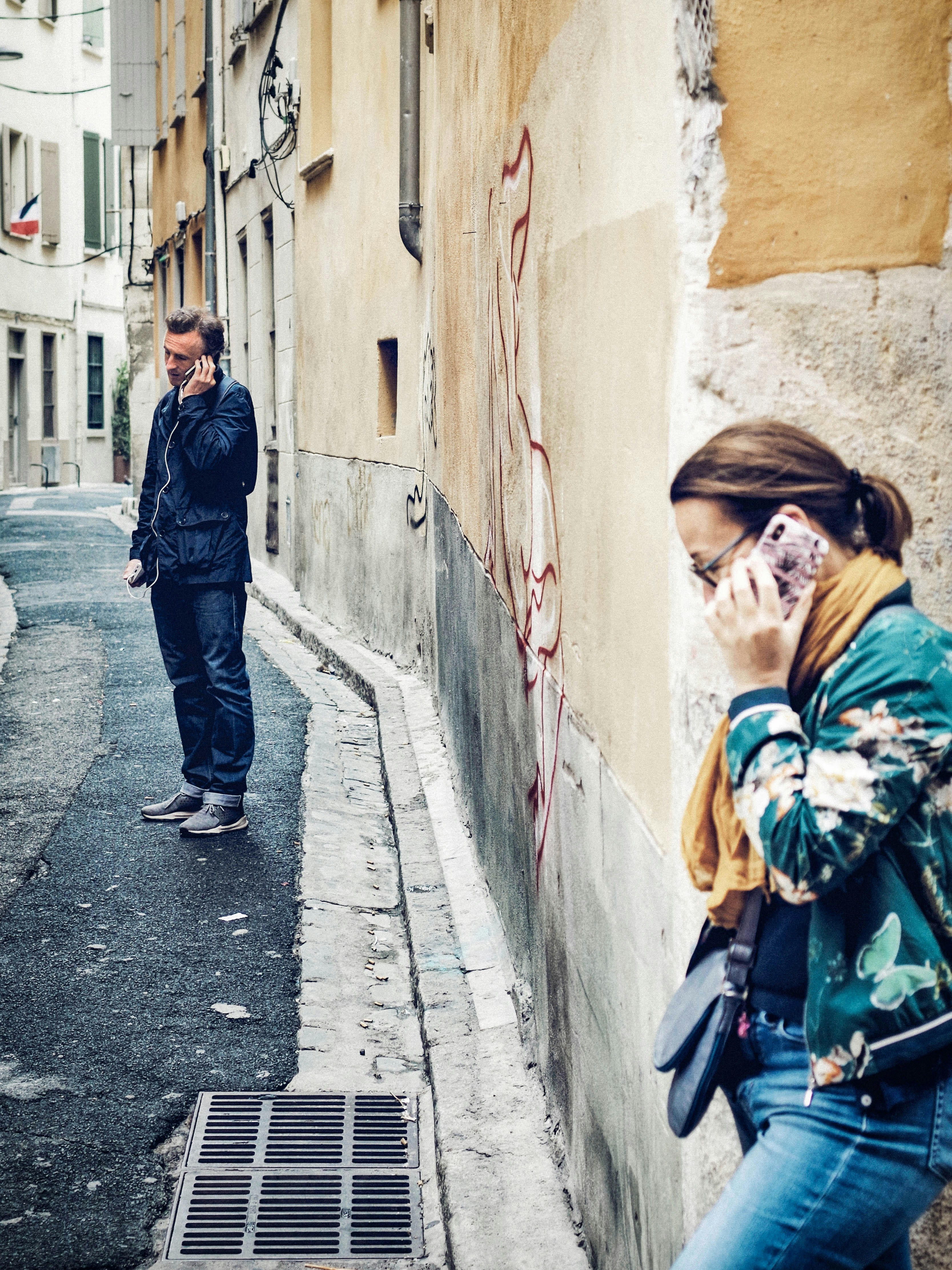 Two people on phone calls in a narrow urban street, each facing opposite directions.