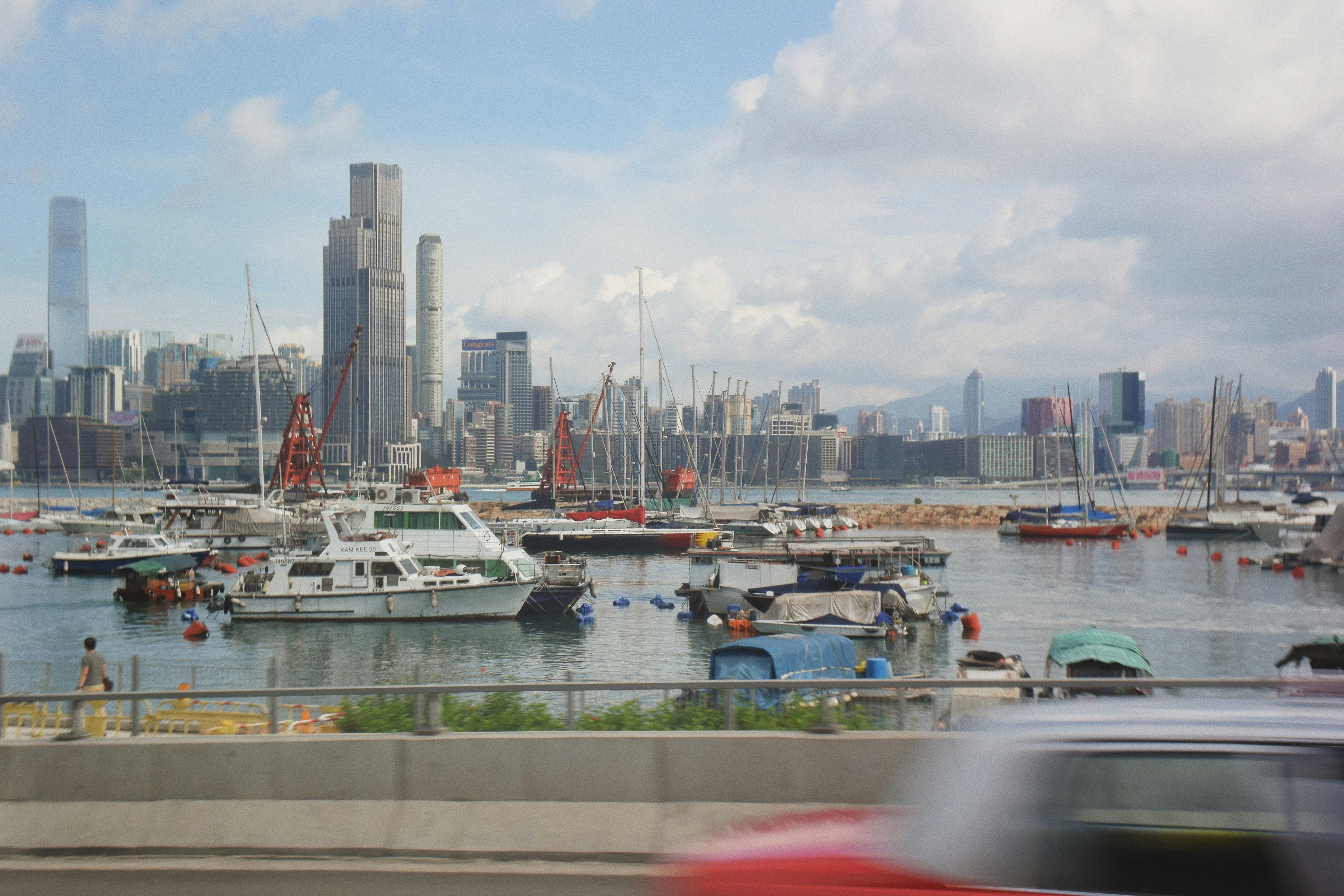 Harbor scene with sailboats and yachts anchored in calm water, a blurred roadway crossing the foreground, and a distant city skyline.