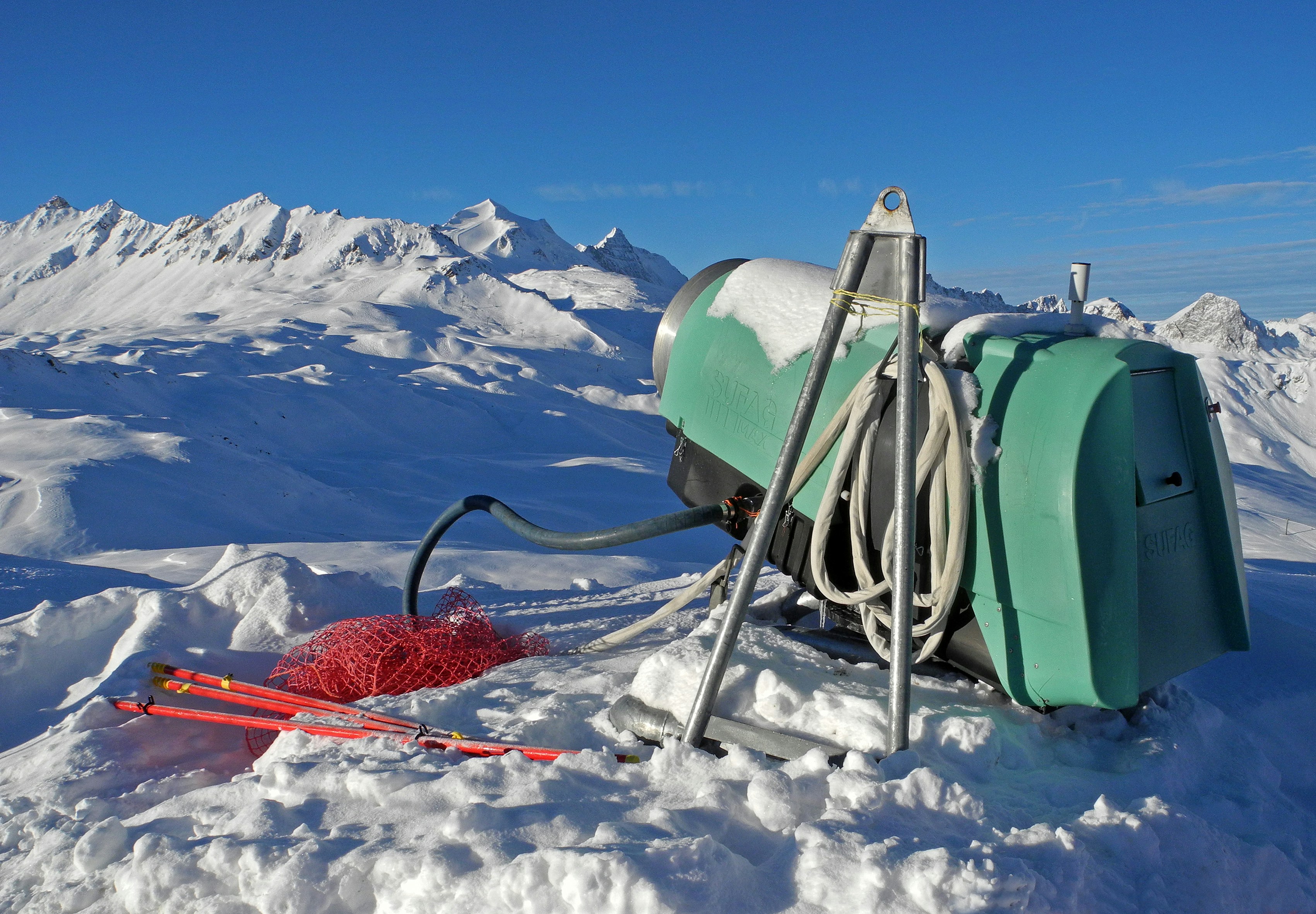 A snow-making machine positioned atop a mountain, surrounded by pristine white snow and distant peaks under a clear blue sky.