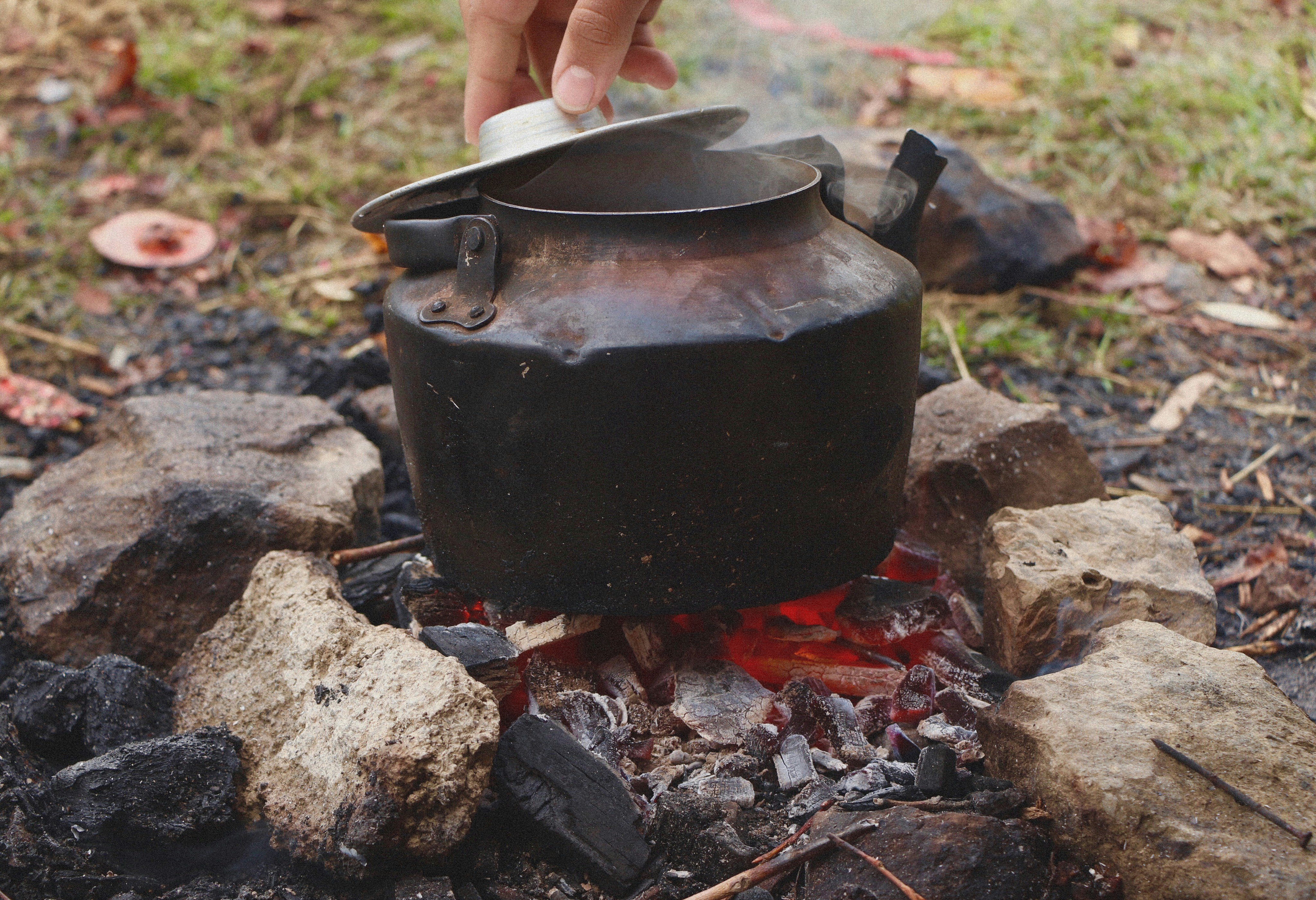 A hand lifts the lid off a black kettle resting on glowing embers, surrounded by rugged stones. The scene evokes the warmth of outdoor cooking.