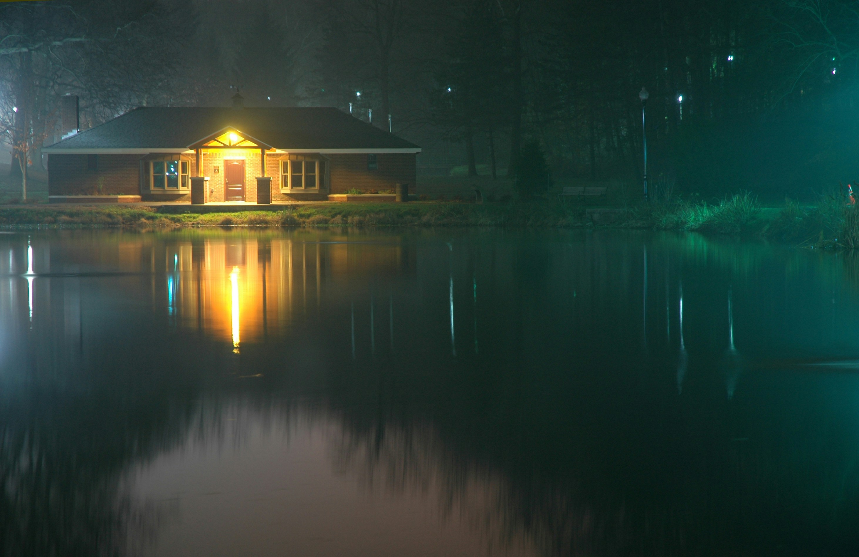 Night photograph of a lamp-lit cabin by a lake, with warm light reflecting on calm water and surrounding trees.