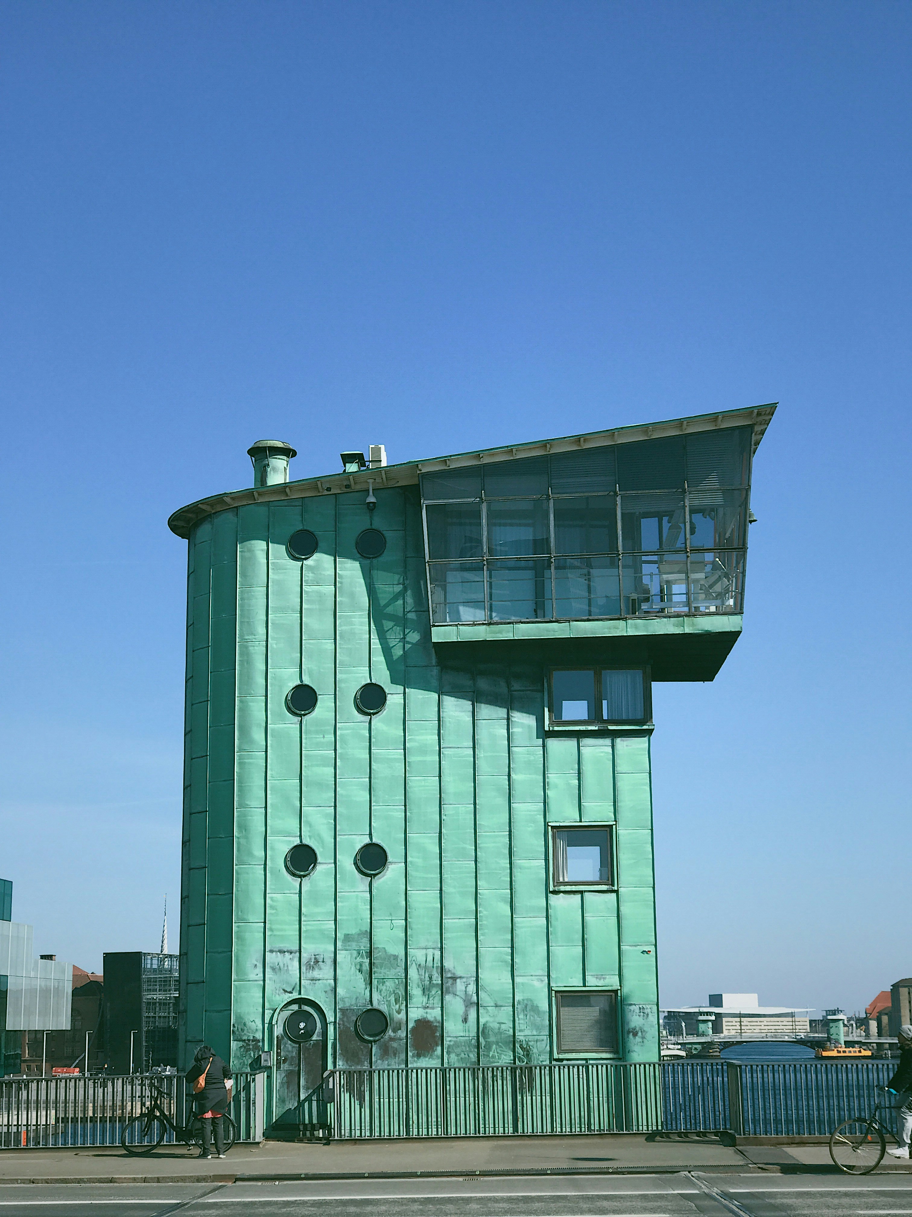 Unique green building with circular windows and a cantilevered upper section, set against a clear blue sky.