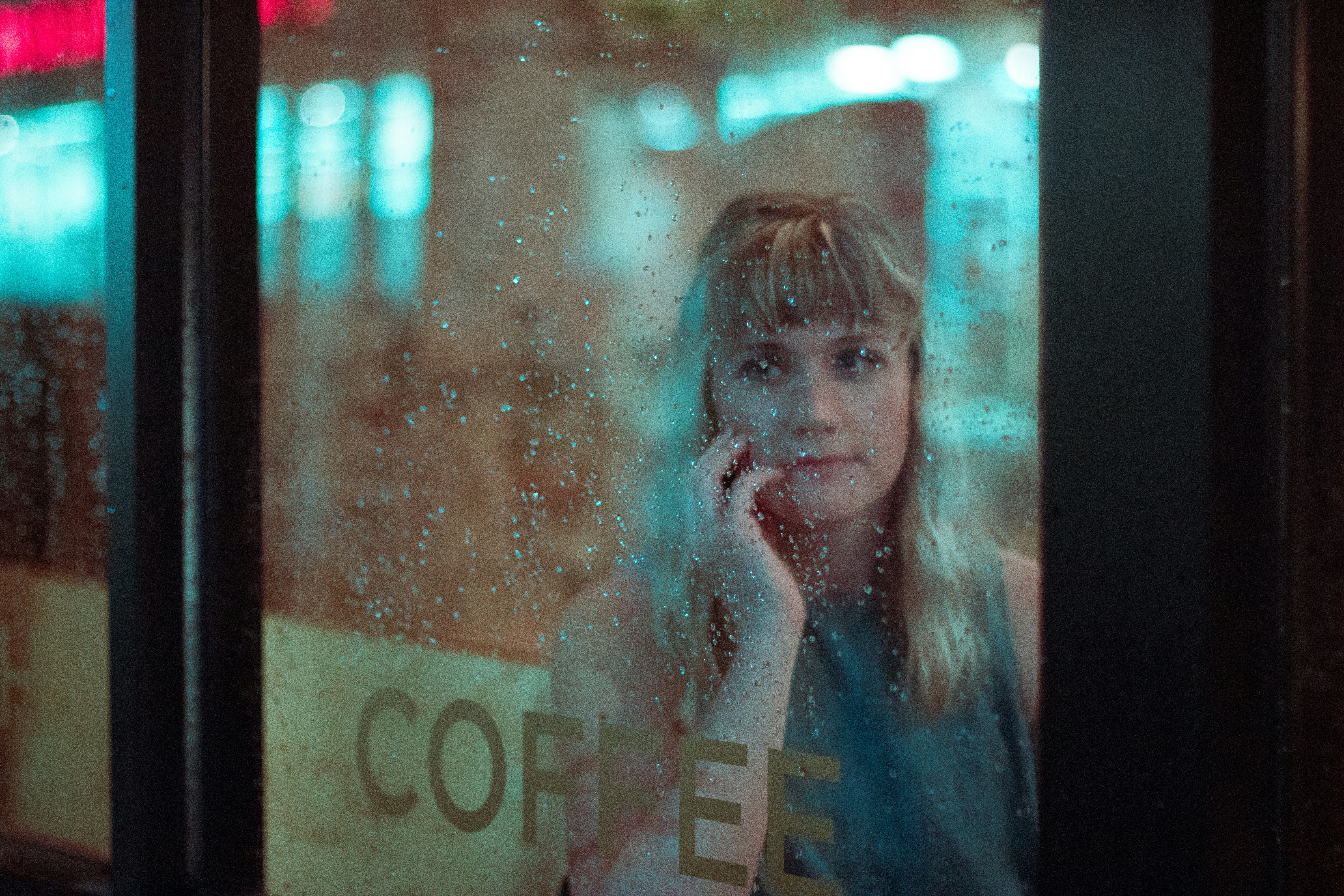 A woman gazes pensively through a rain-speckled window, framed by the warm glow of a coffee shop sign. The atmosphere evokes a sense of introspection and quiet contemplation.