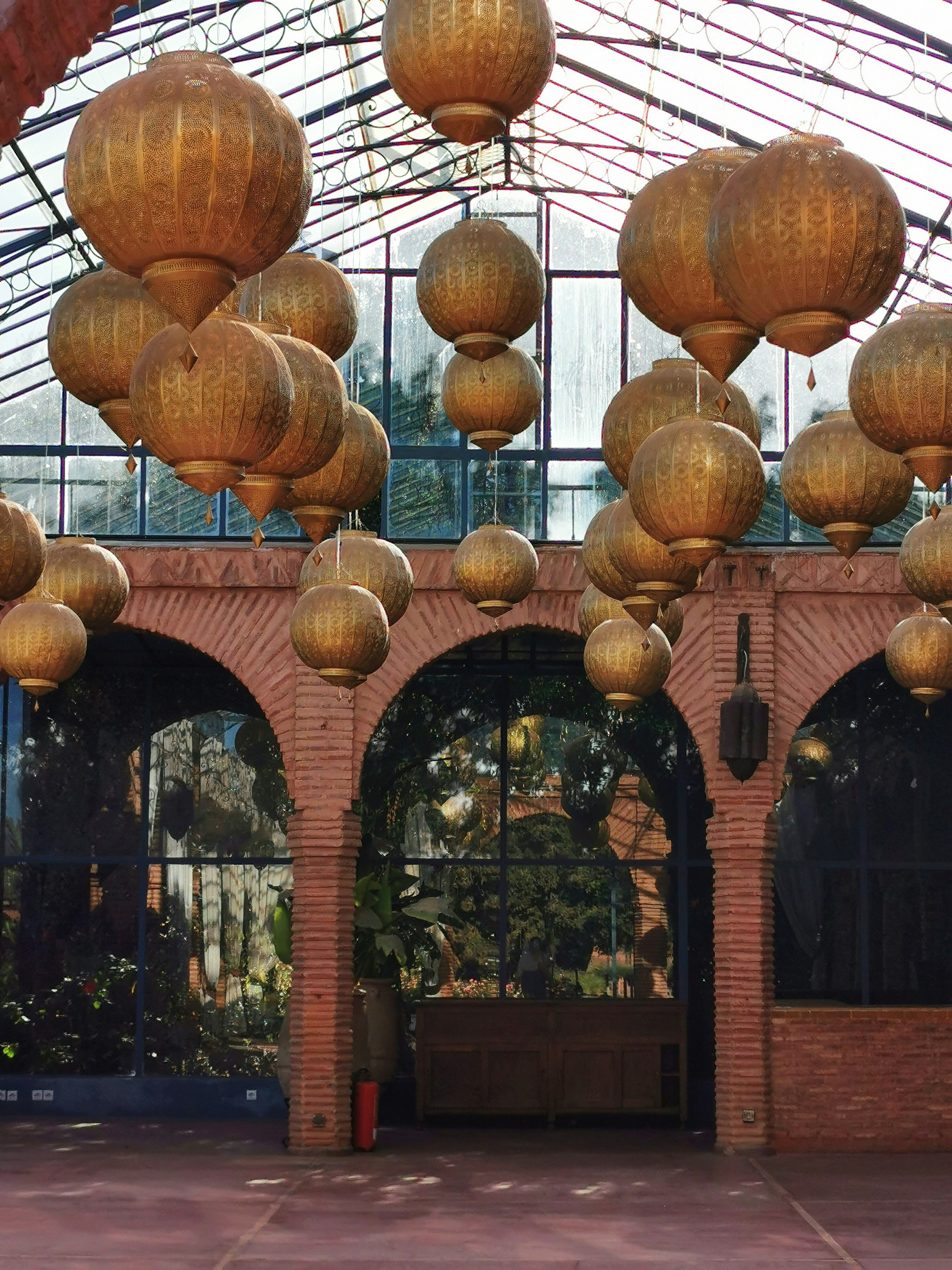 A collection of ornate golden lanterns suspended from a glass ceiling, illuminating a brick-walled atrium filled with greenery.