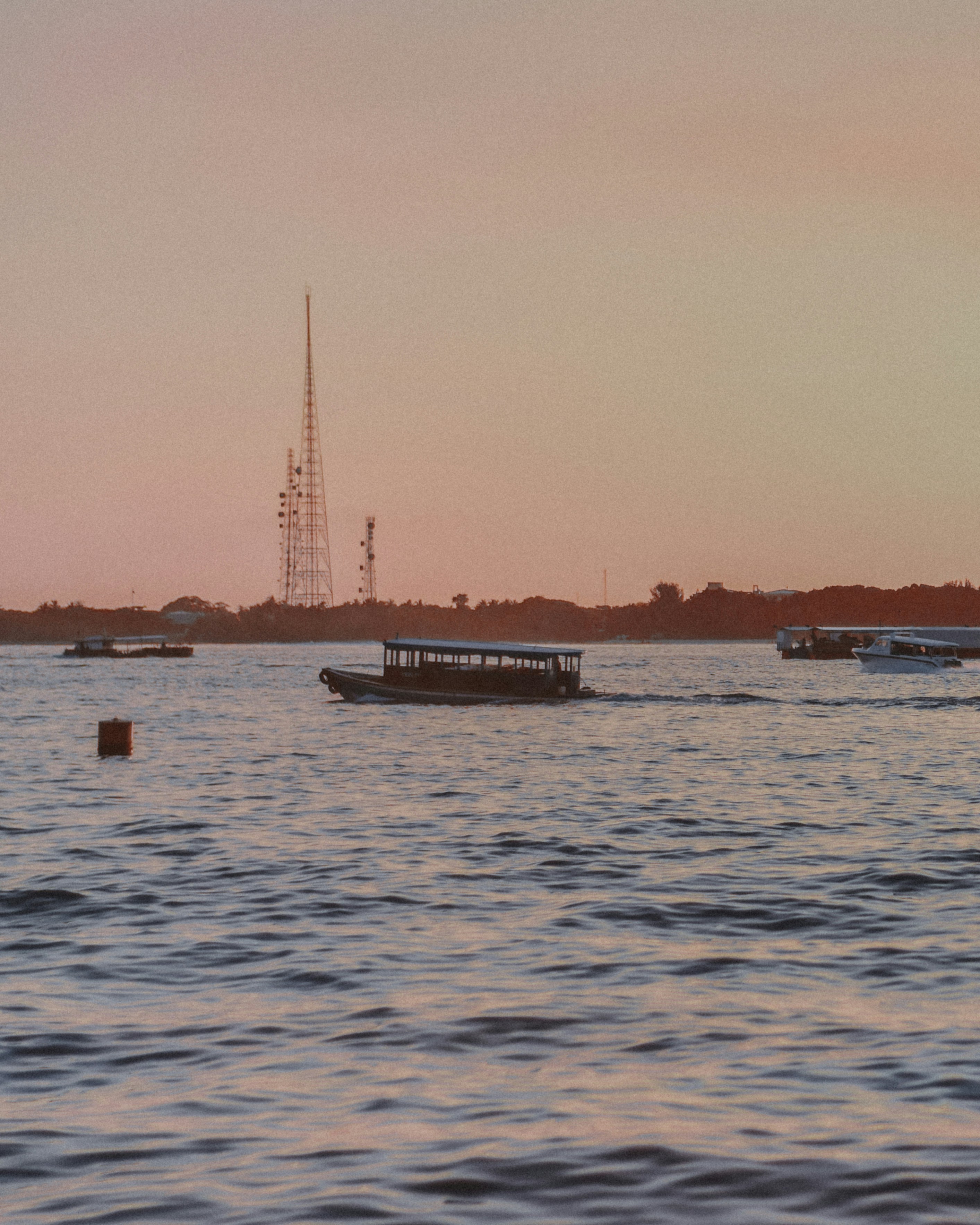 A boat glides across a tranquil river at dusk, with a silhouette of a radio tower in the background. Soft pastel hues blend in the sky.