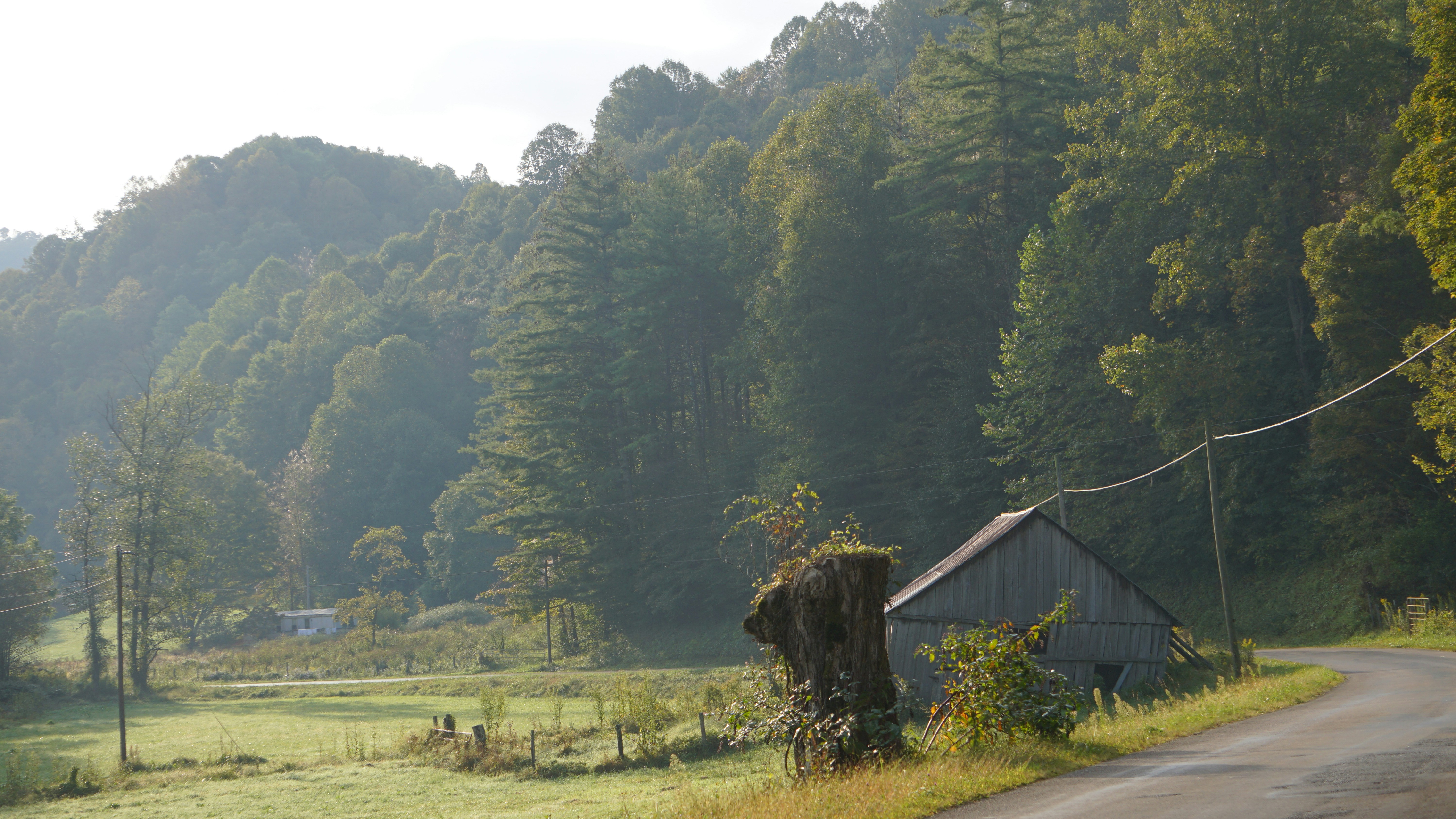 A quaint wooden barn nestled beside a winding road, surrounded by lush green hills and misty trees in the early morning light.