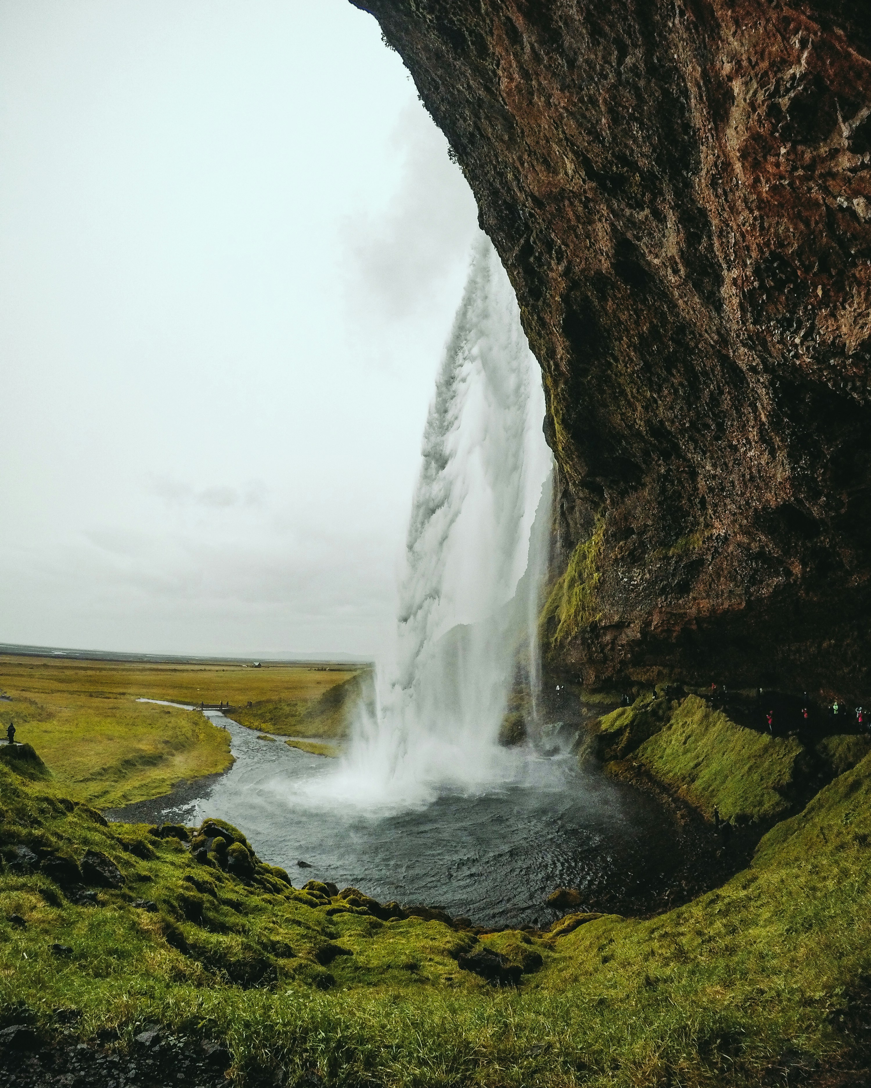 Water cascades dramatically from a rocky cliff, creating a misty curtain over a lush green landscape. The scene captures the dynamic interaction between land and water.