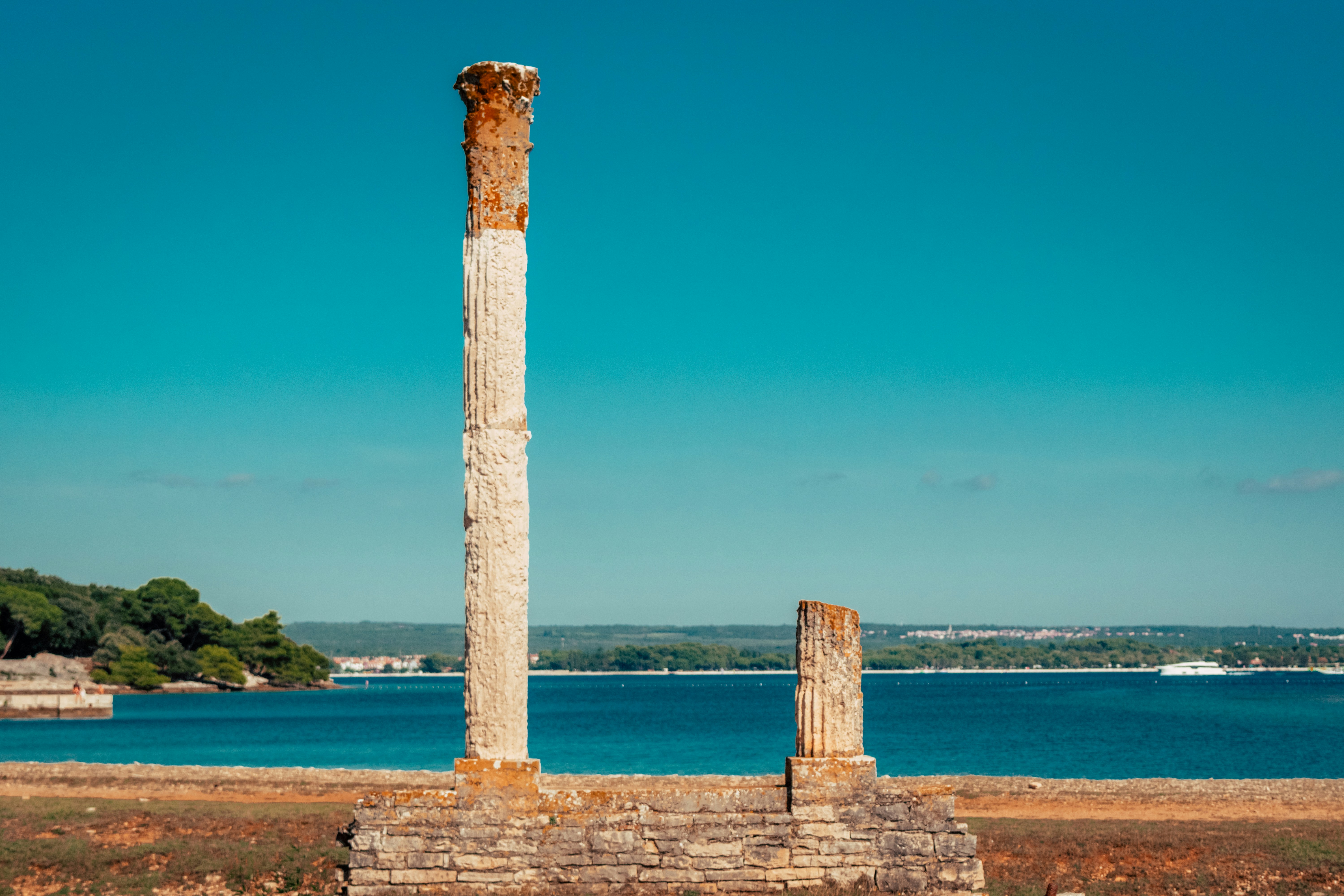 Two weathered columns stand resiliently by the water's edge, framed by a tranquil blue sky and distant landscape.