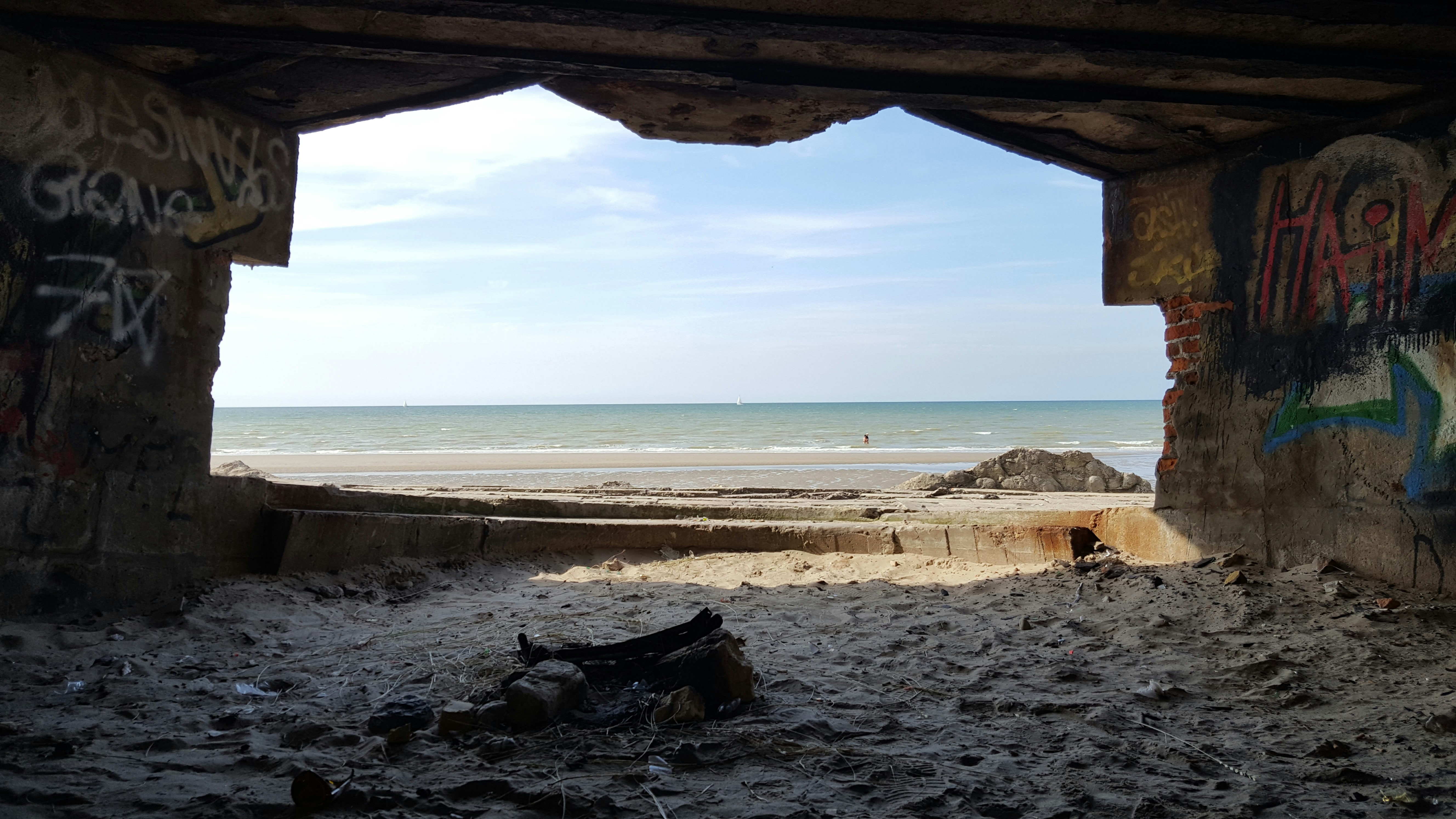 View of a tranquil beach framed by the ruins of an old structure, with graffiti adding a touch of urban contrast. The calm sea stretches into the horizon.