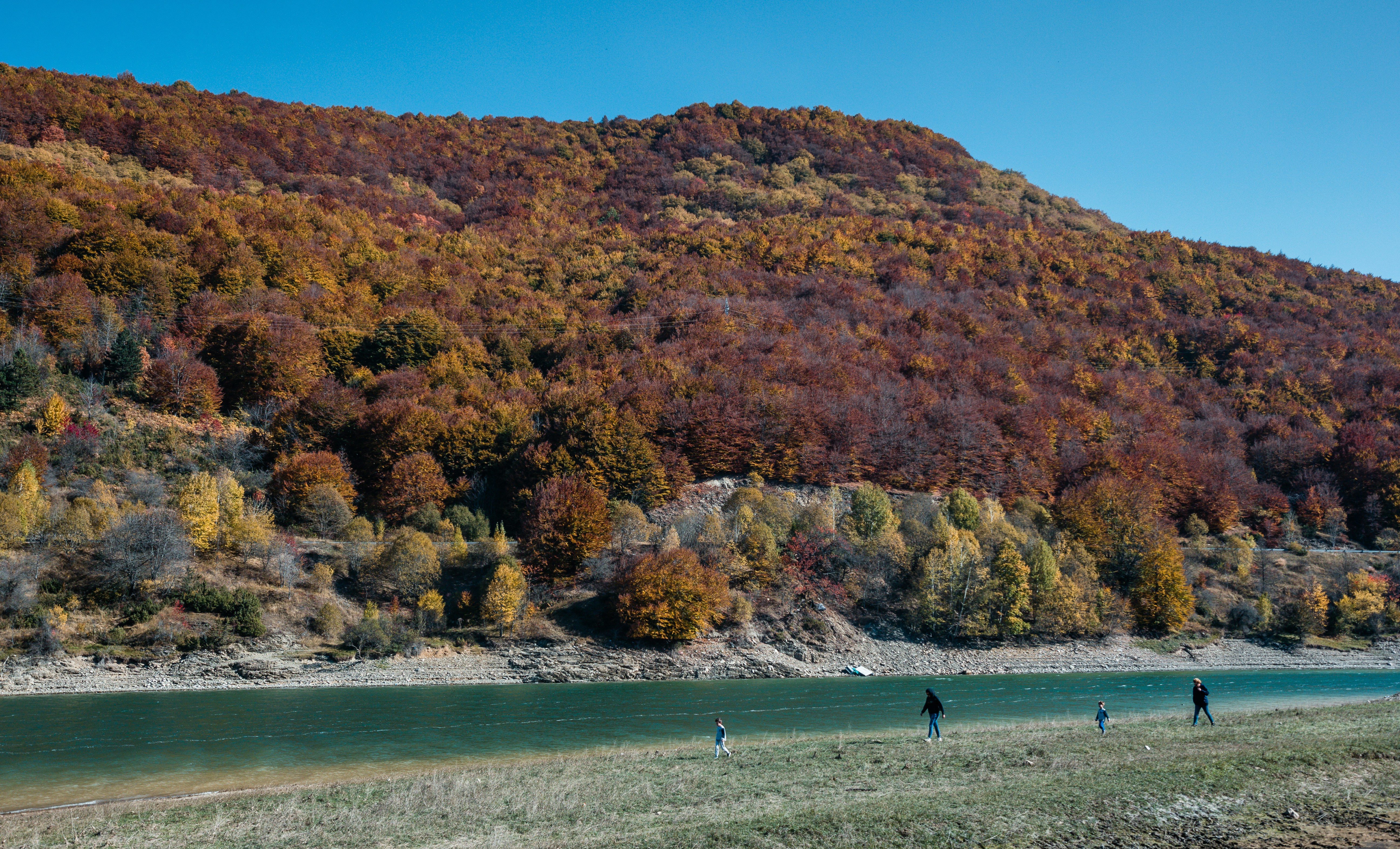 Families stroll along the riverbank, surrounded by vibrant autumn foliage reflecting in the water. A serene landscape captures the essence of fall.