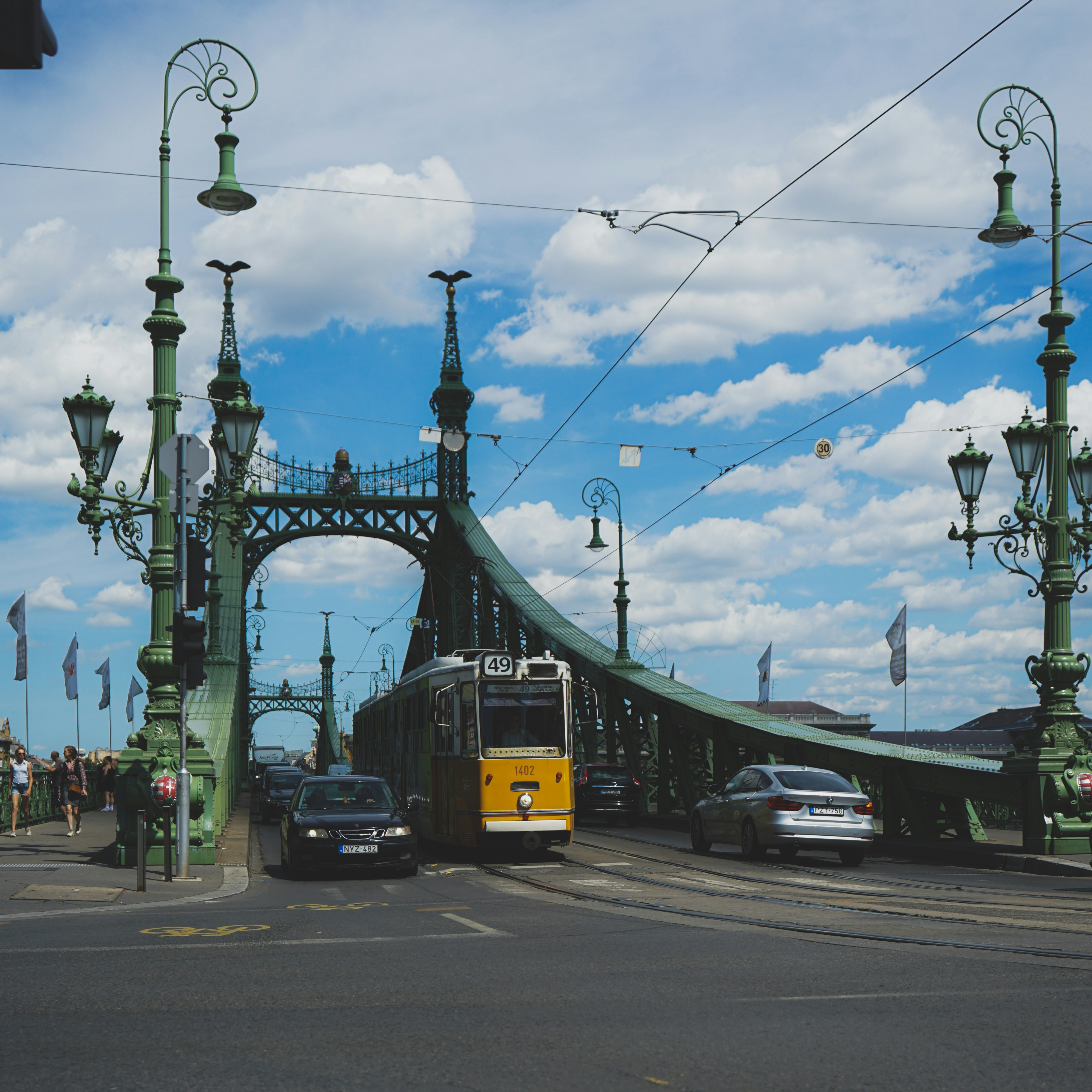 Tram crossing a vibrant green bridge under a bright blue sky with fluffy clouds, surrounded by historic architecture and bustling city life.