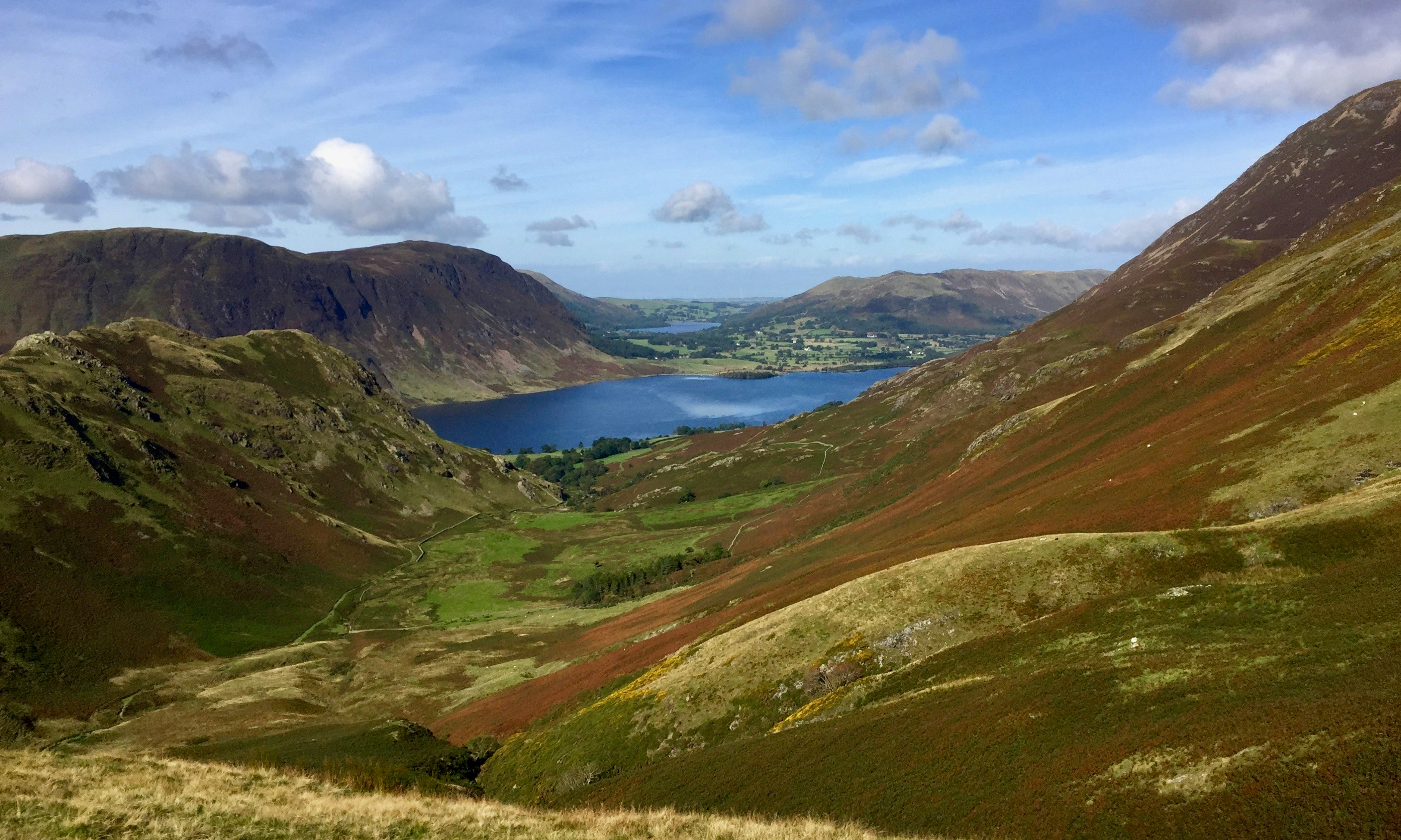 Vibrant green valleys meet a serene blue lake, framed by rugged hills under a partly cloudy sky.