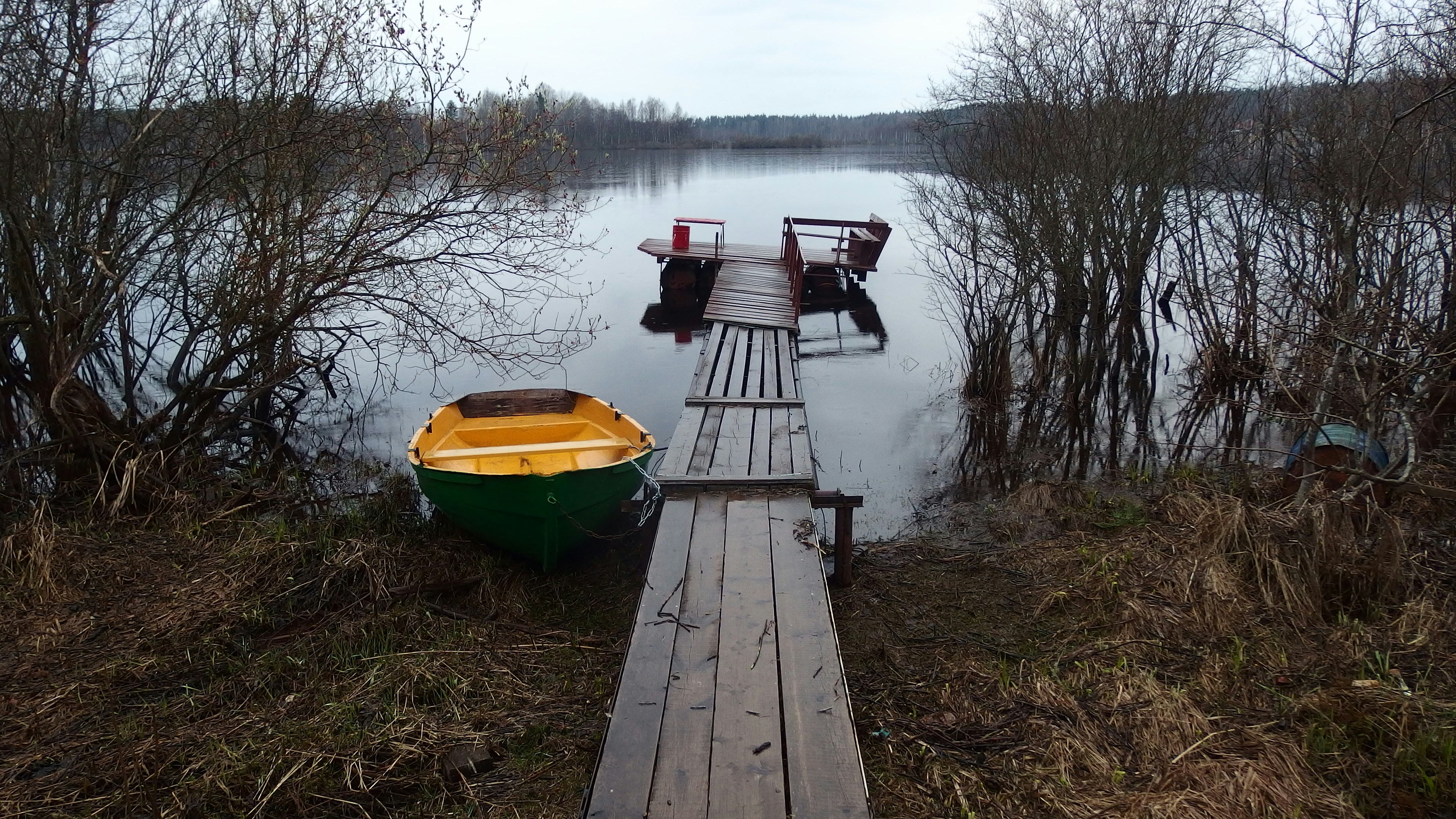 Wooden dock extending into a calm lake, flanked by a green and yellow boat and bare trees. The tranquil water reflects the overcast sky.