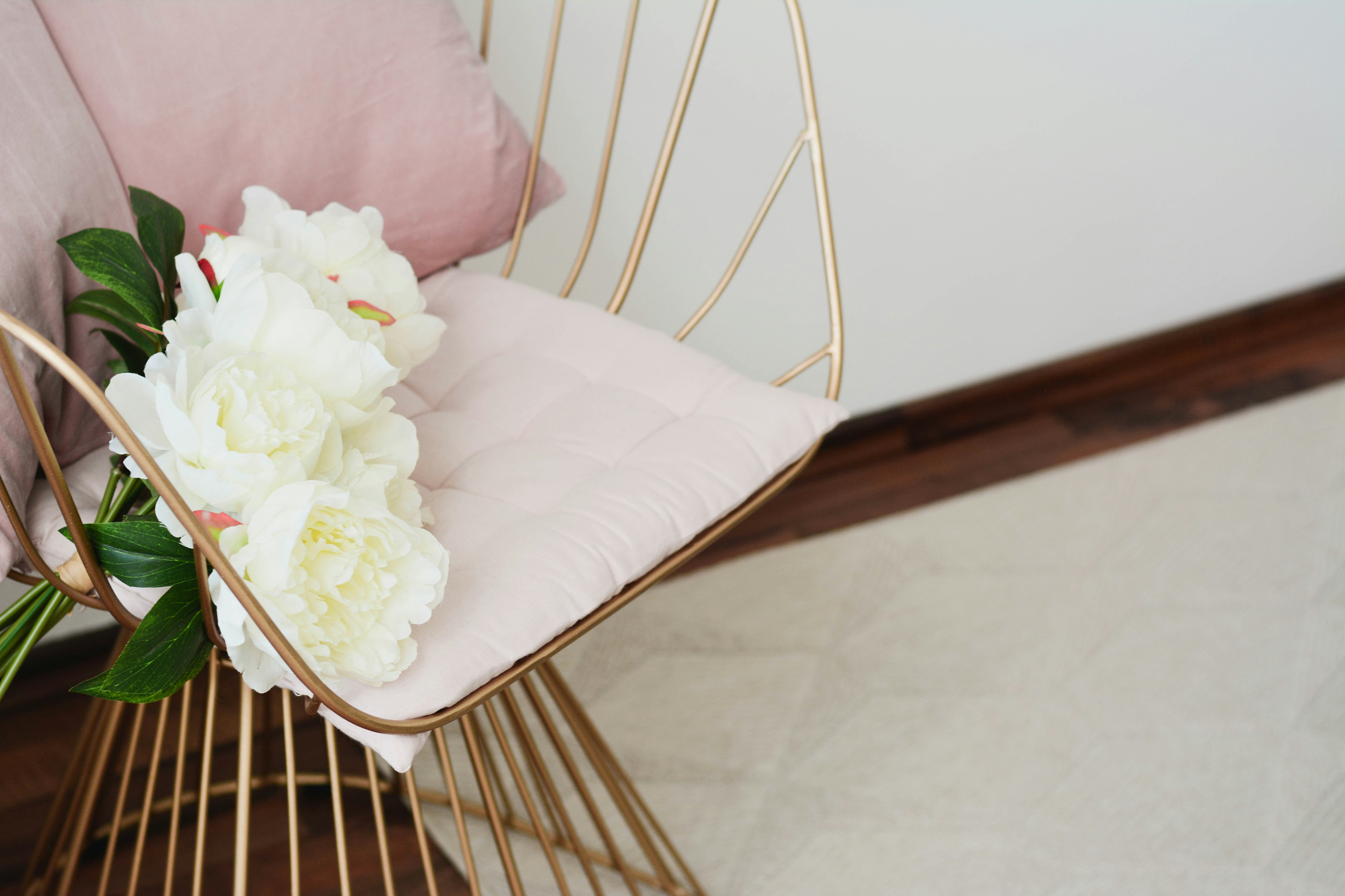 White peonies rest gracefully on a pastel pink cushion of a modern wire chair, accentuating a serene interior setting.