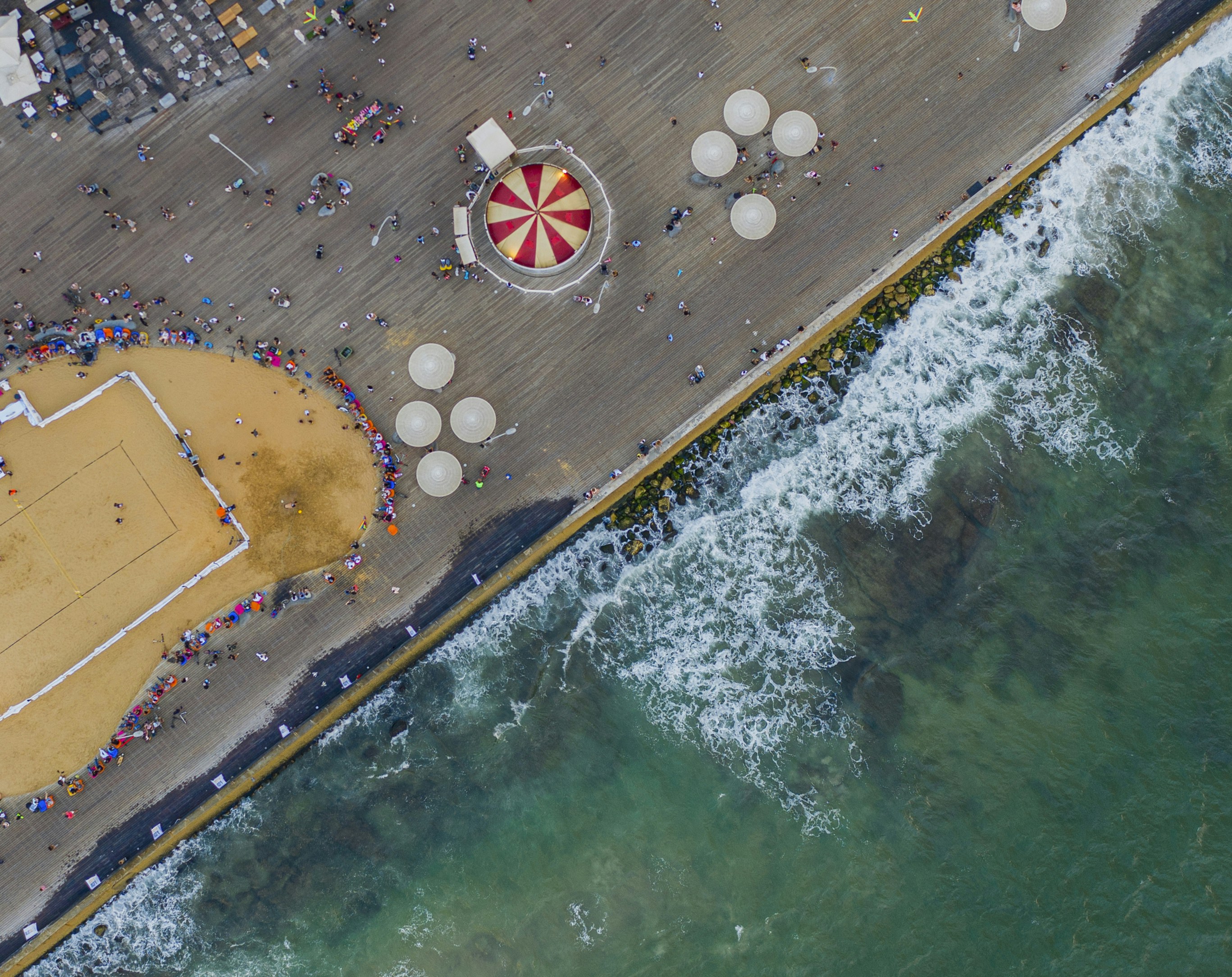 Aerial view of a vibrant seaside carnival featuring a colorful carousel, beachgoers, and crashing waves along the shore.