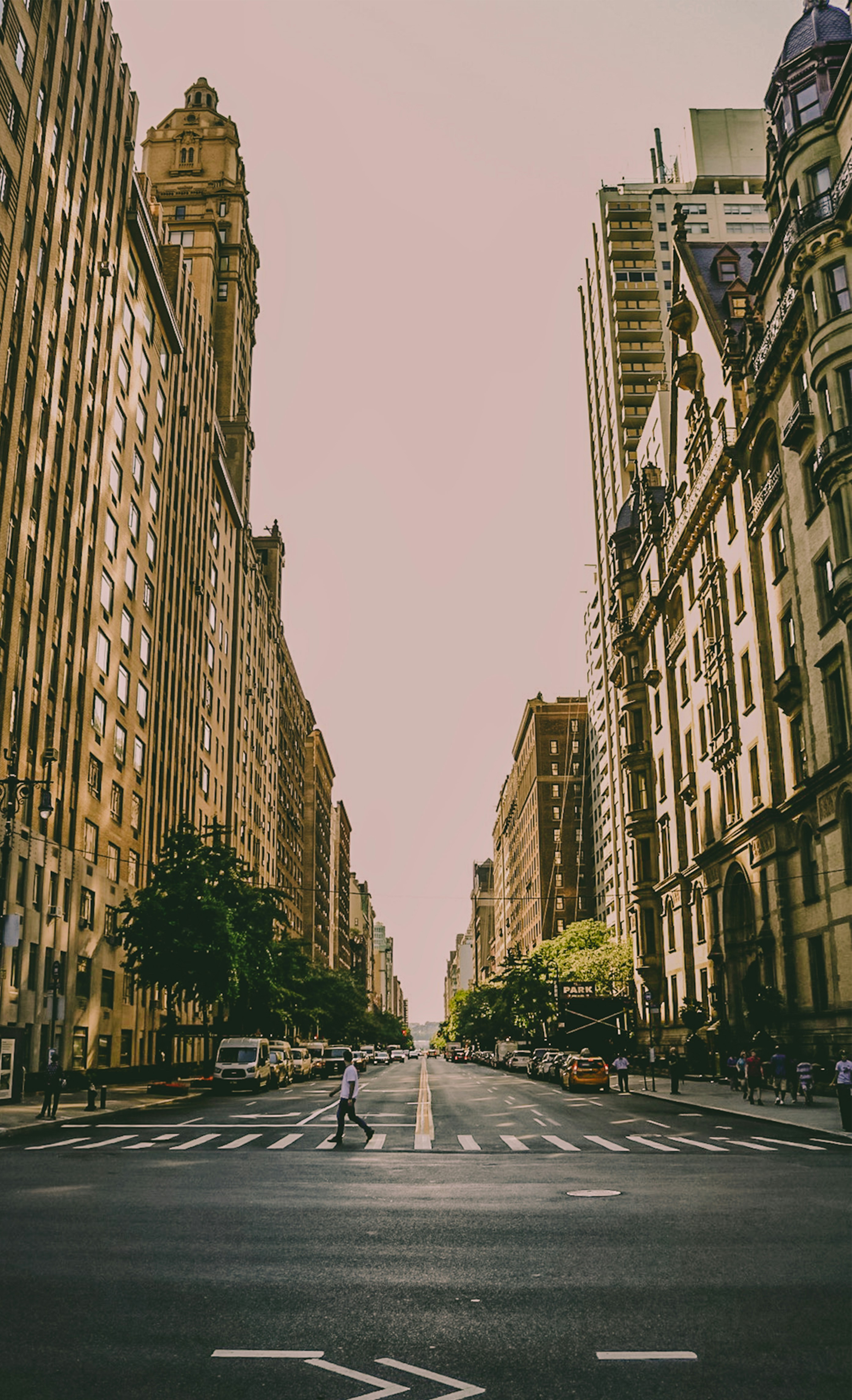 A busy city street lined with towering buildings, capturing the essence of urban life as pedestrians navigate the crosswalk under a softly glowing sky.