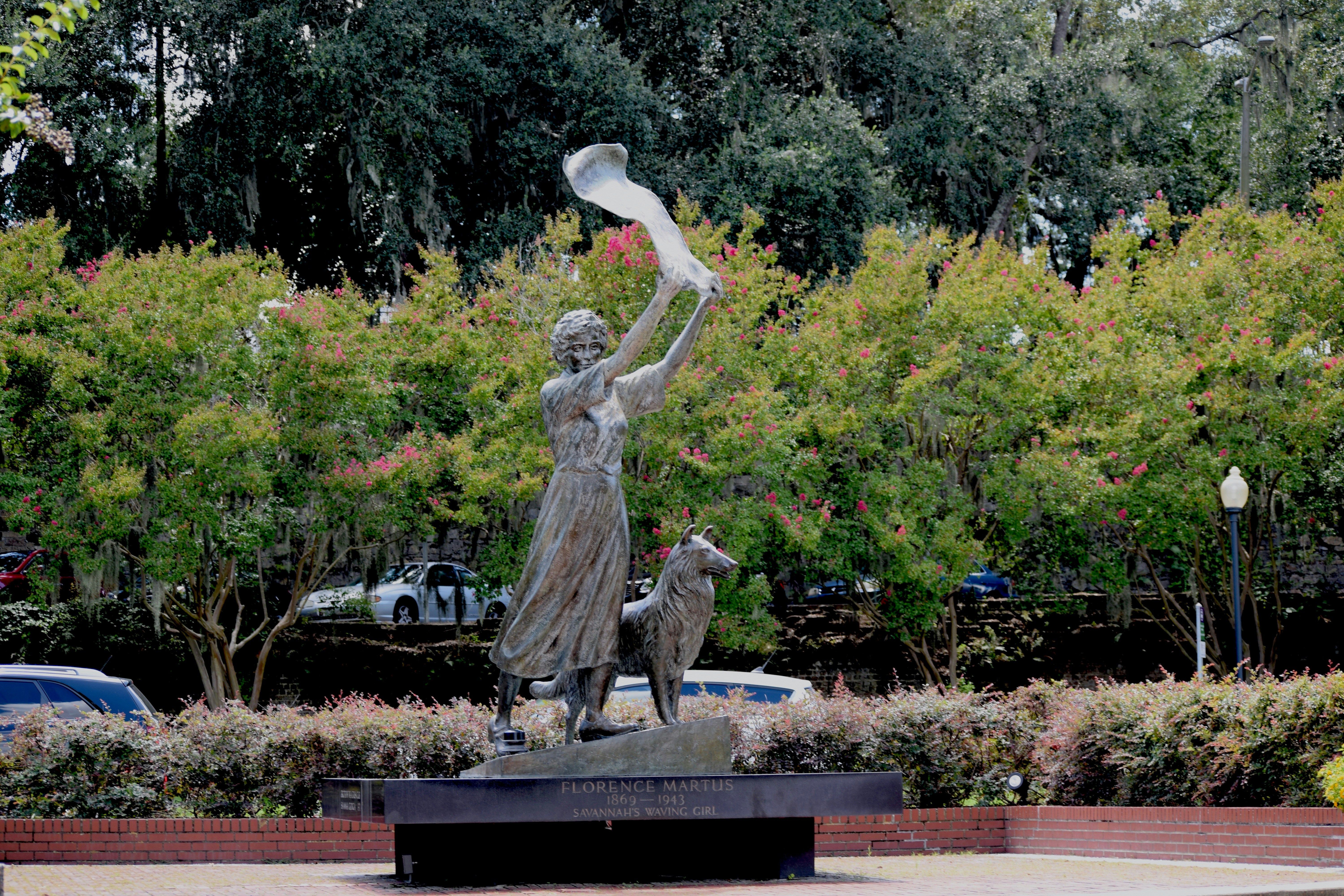 Bronze statue of a woman raising a scarf, set against vibrant autumn foliage in a park.