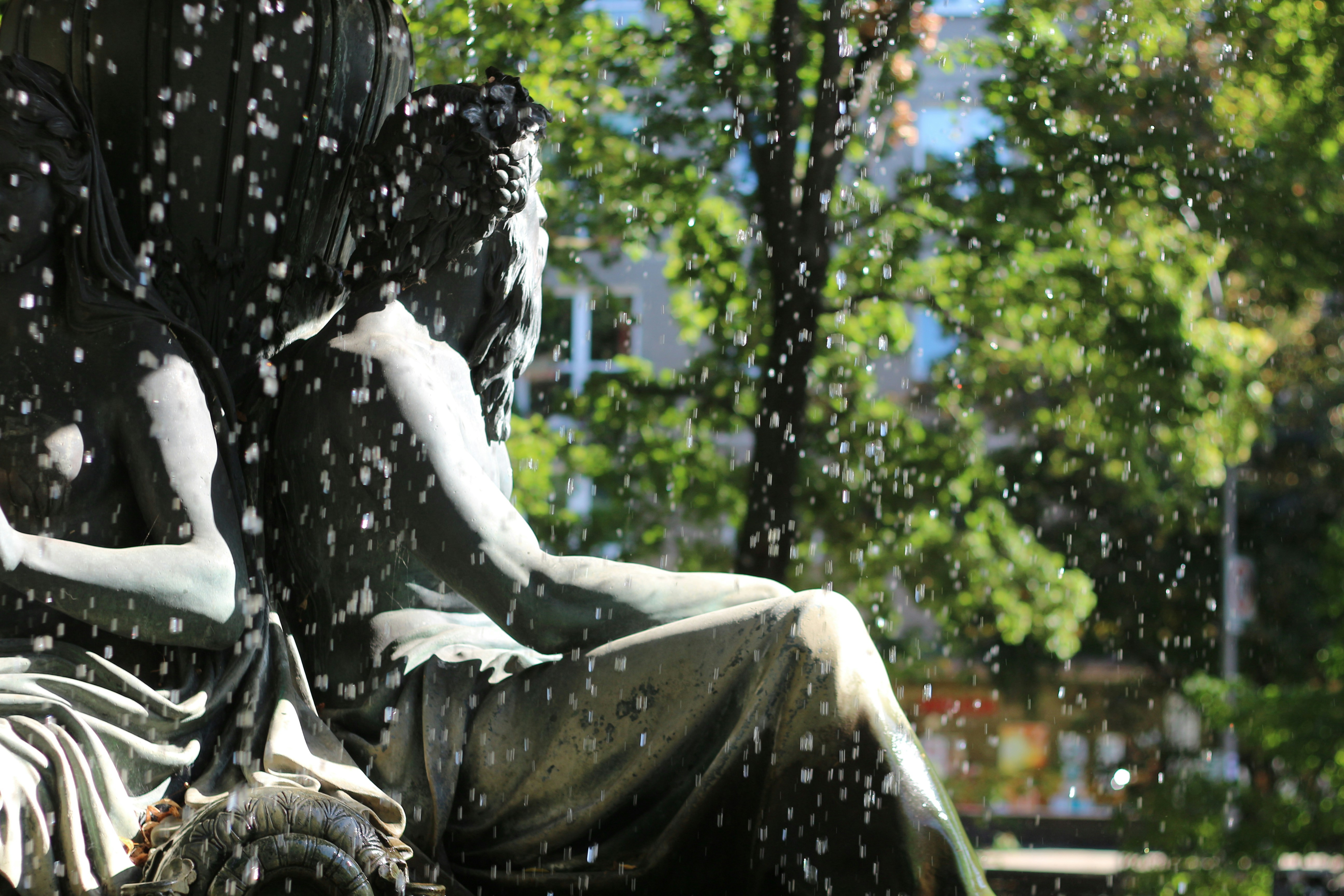 Photograph of a dark marble statue in a fountain, droplets cascading across its surface as sunlight filters through green trees.