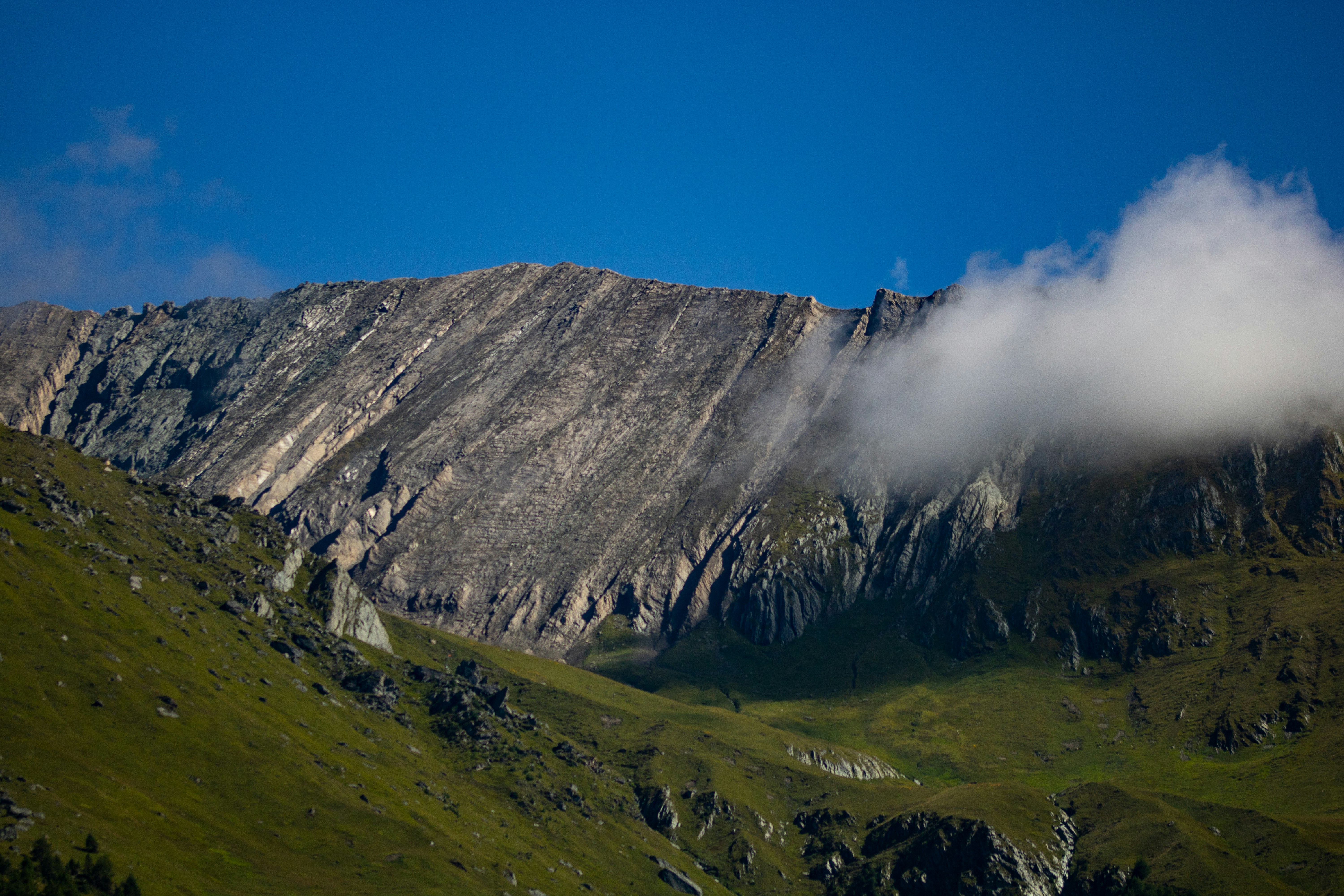 A dramatic mountain ridge under a clear blue sky, with a cloud gently resting on its peak. The natural contours and textures of the rock face are prominently displayed.