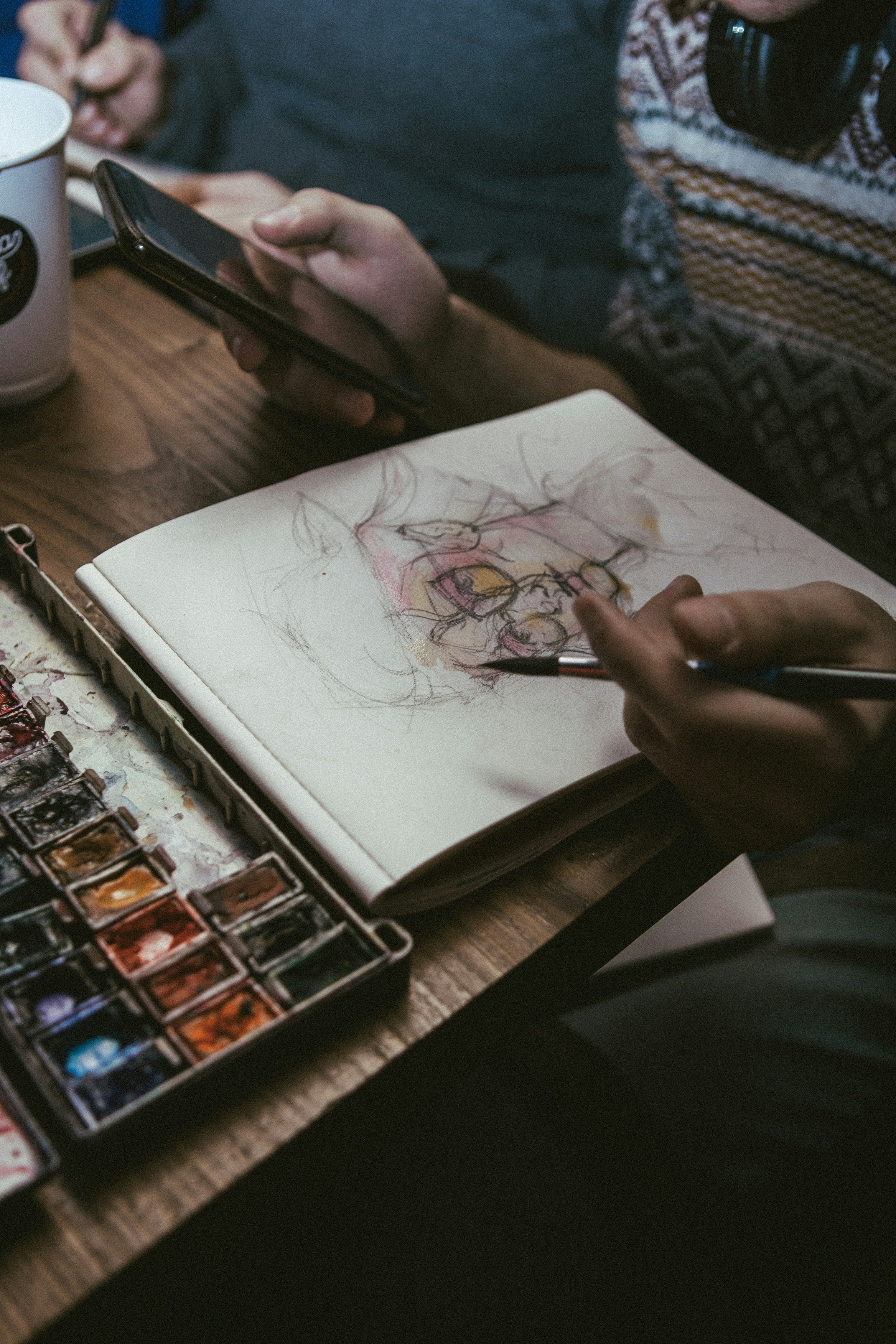 A hand sketches a vibrant illustration in a notebook while a palette of watercolors rests nearby, capturing the essence of creativity in a cozy setting.