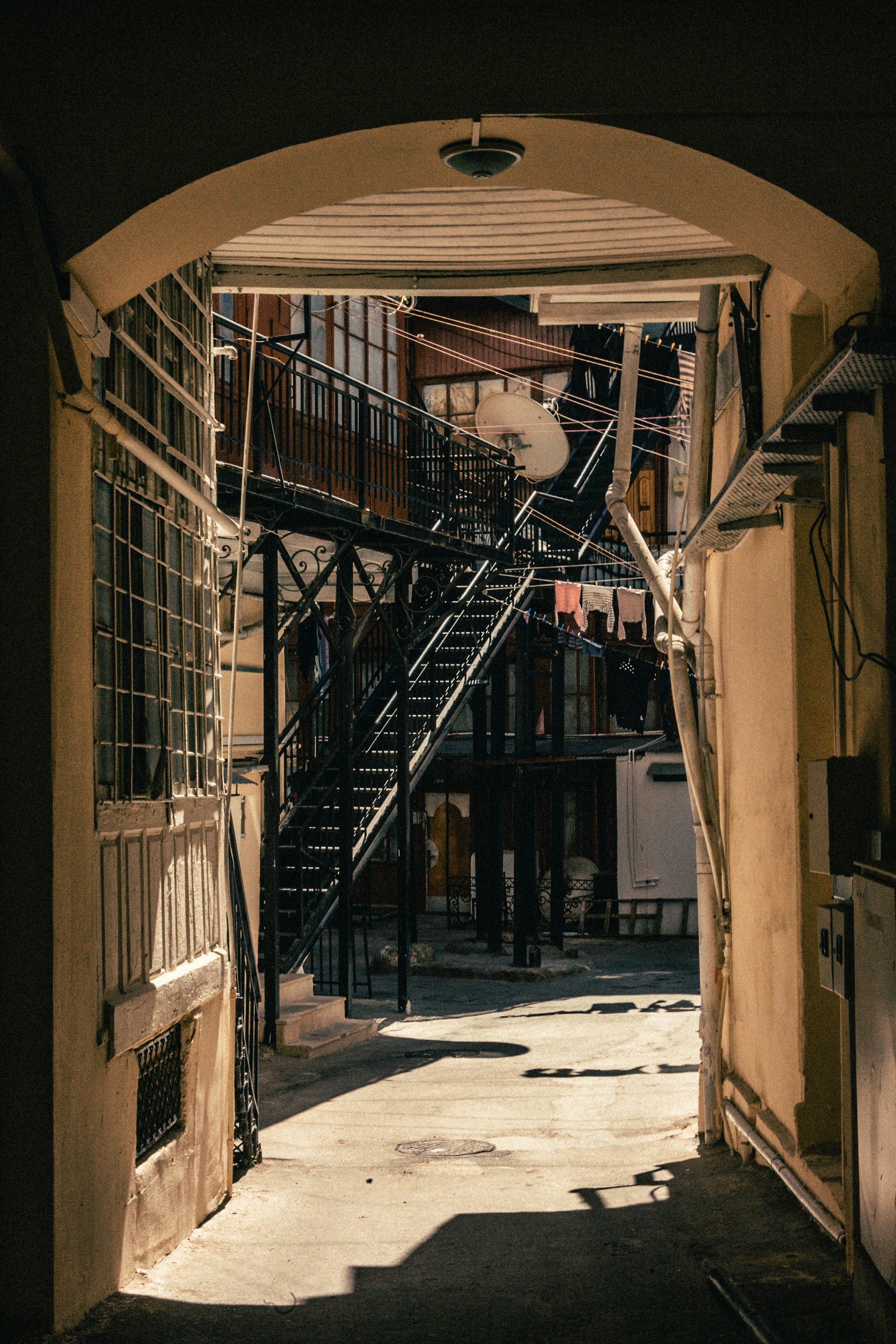 Narrow alleyway with a staircase and hanging laundry, framed by an archway. Sunlight casts shadows on the ground.