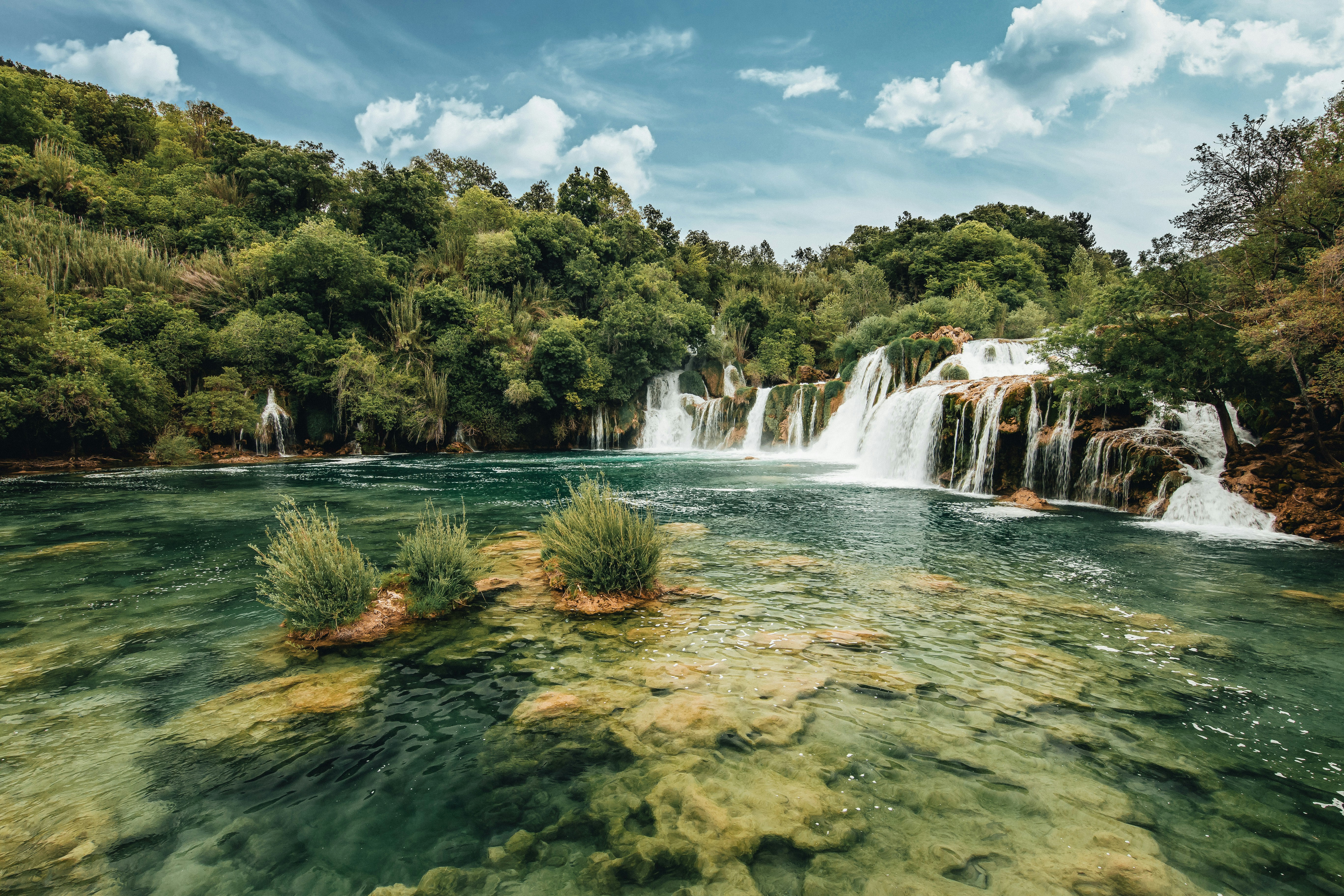 Lush green landscape surrounding a tranquil waterfall, with clear water reflecting the sky above. The scene captures the harmony of nature.