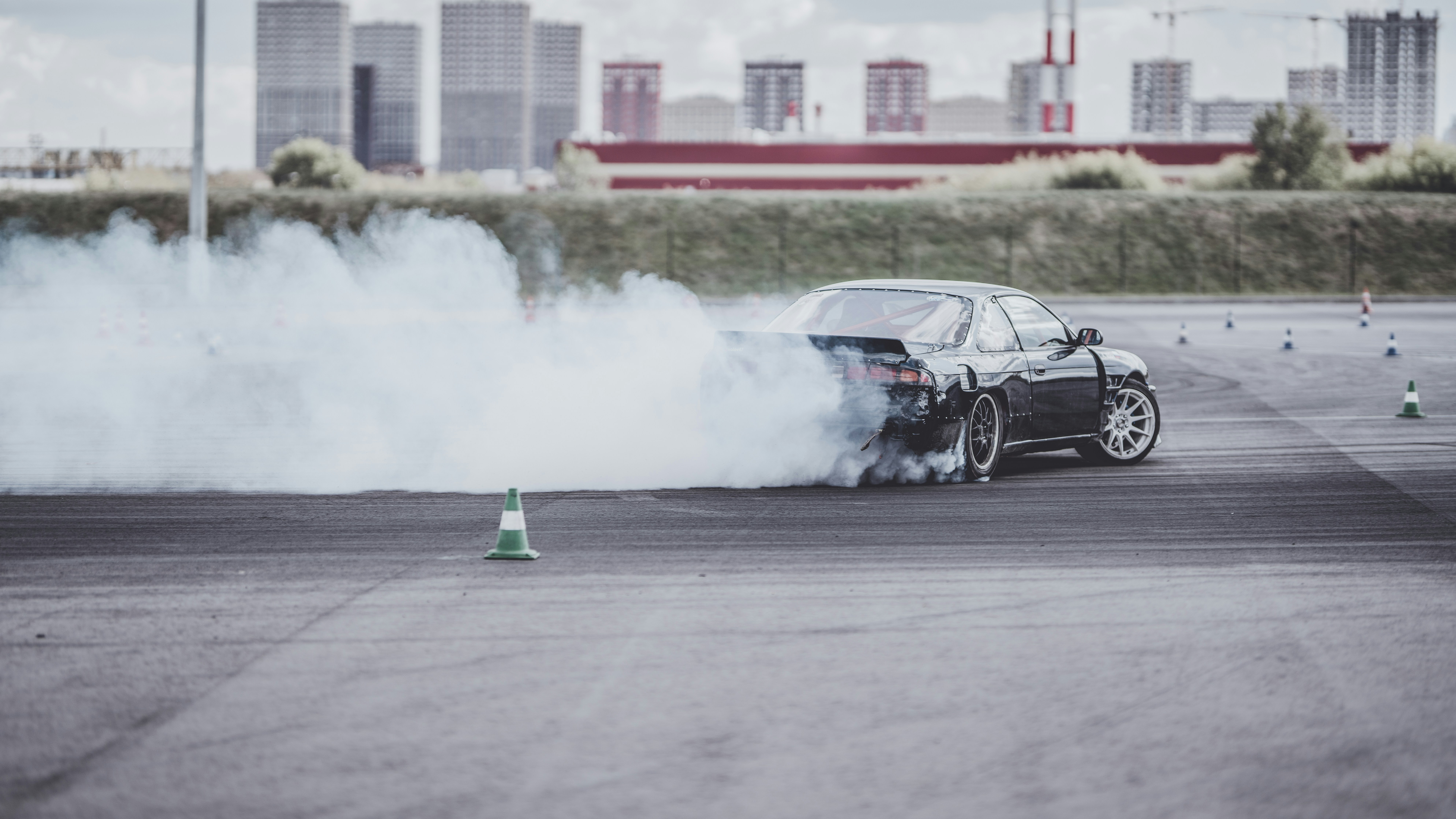 Black car executing a drift maneuver, surrounded by tire smoke on an asphalt track with distant city buildings.