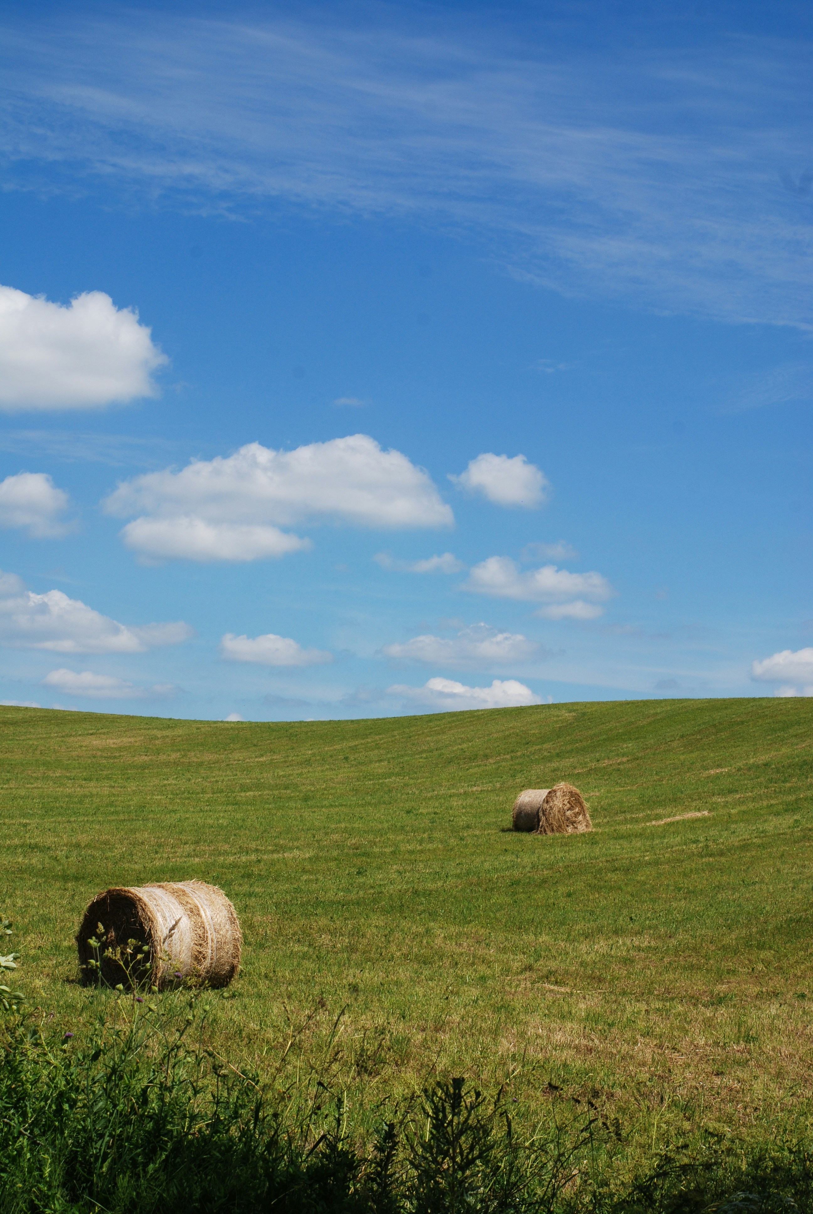 Two hay bales resting on a lush green field under a bright blue sky dotted with clouds.
