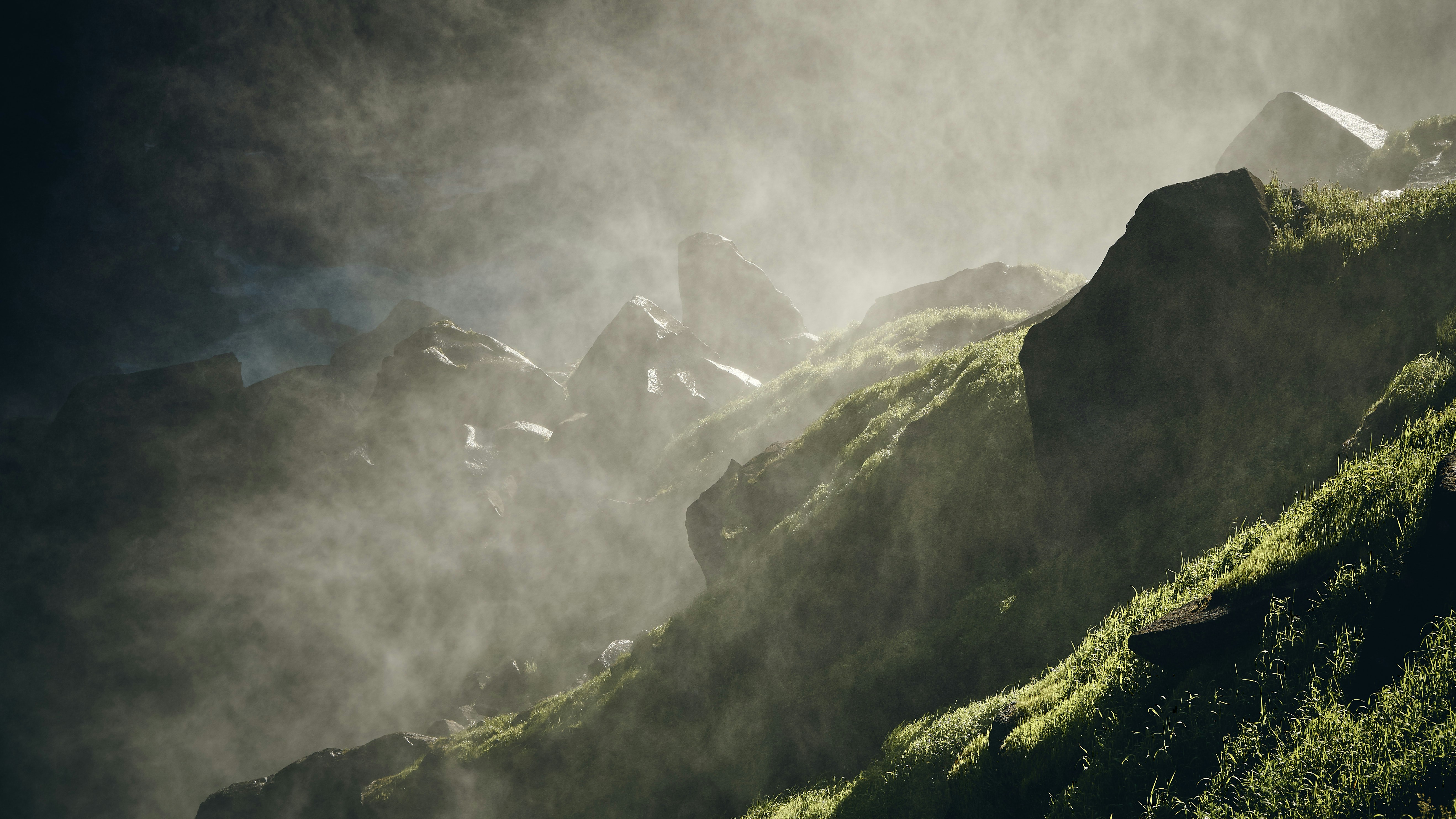This captivating image captures a rocky hillside enveloped in a soft mist, creating an ethereal atmosphere. The composition highlights the contrast between the rugged, shadowed rocks and the vibrant green moss that covers them. The interplay of light and shadow adds depth, while the mist diffuses the sunlight, casting a serene, almost mystical ambiance across the scene.