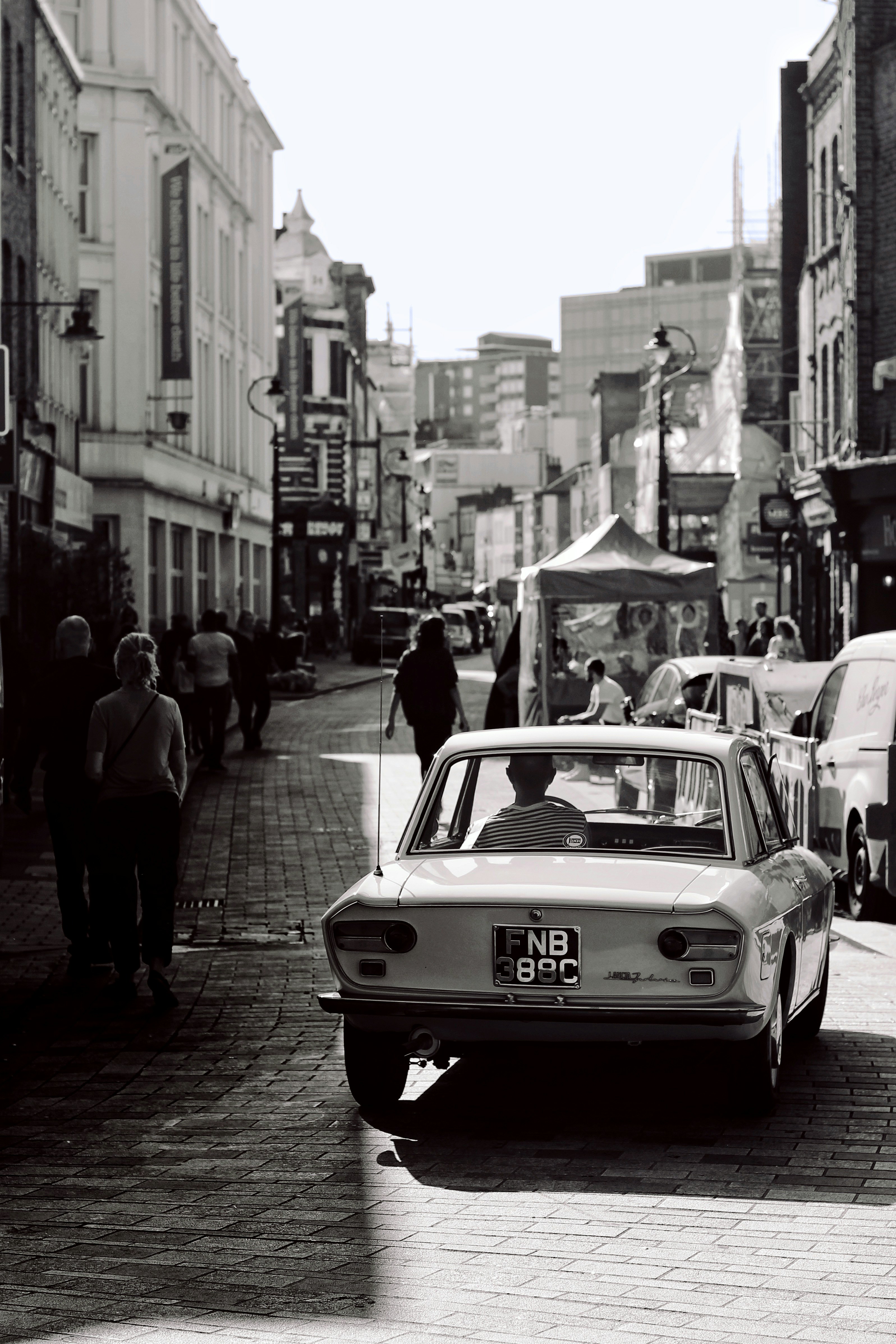 Vintage car parked on a bustling street, surrounded by pedestrians and shops, capturing the essence of city life. The scene is rendered in black and white, emphasizing its timeless quality.