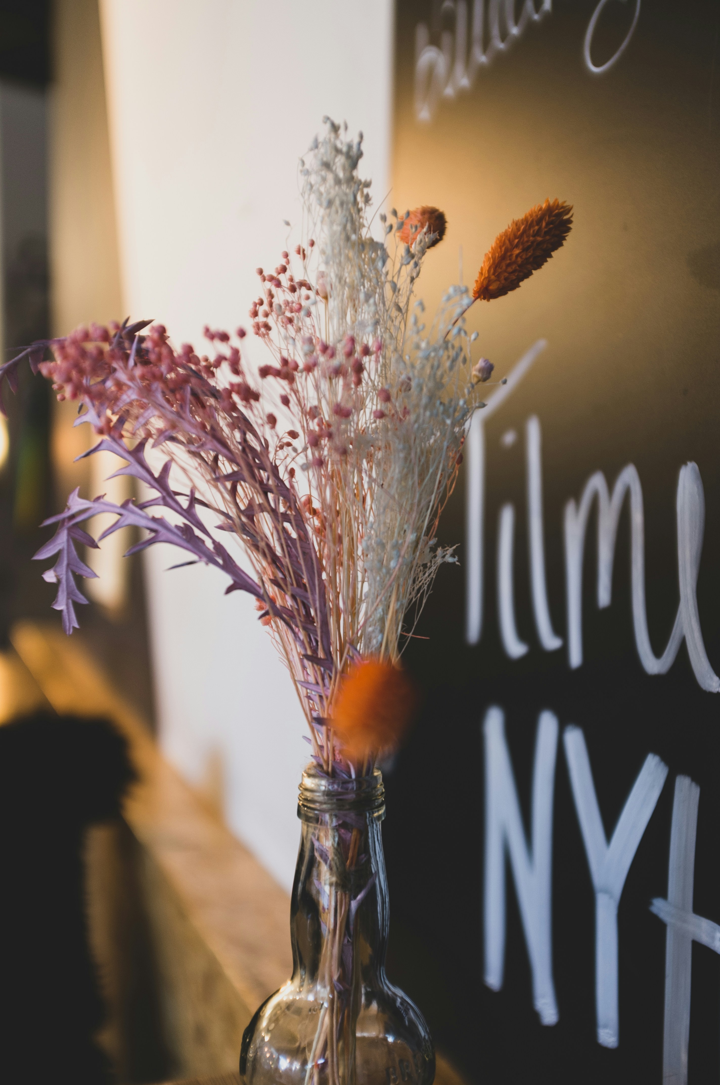Delicate dried flowers arranged in a glass vase against a chalkboard backdrop with handwritten text. The composition highlights the interplay of natural textures and artistic expression.