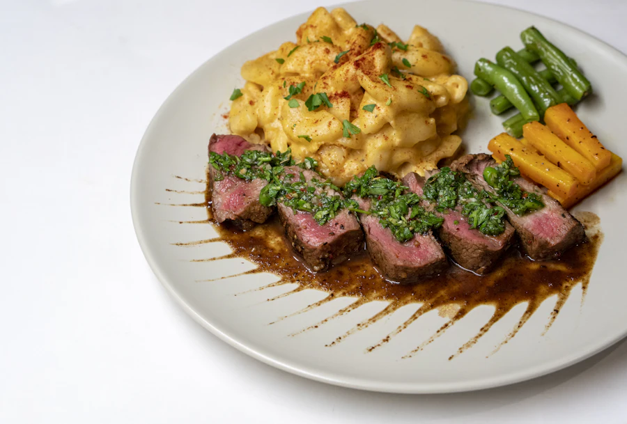 Sliced steak topped with herb sauce on a white plate, served with creamy macaroni and sides of green beans and glazed carrots.
