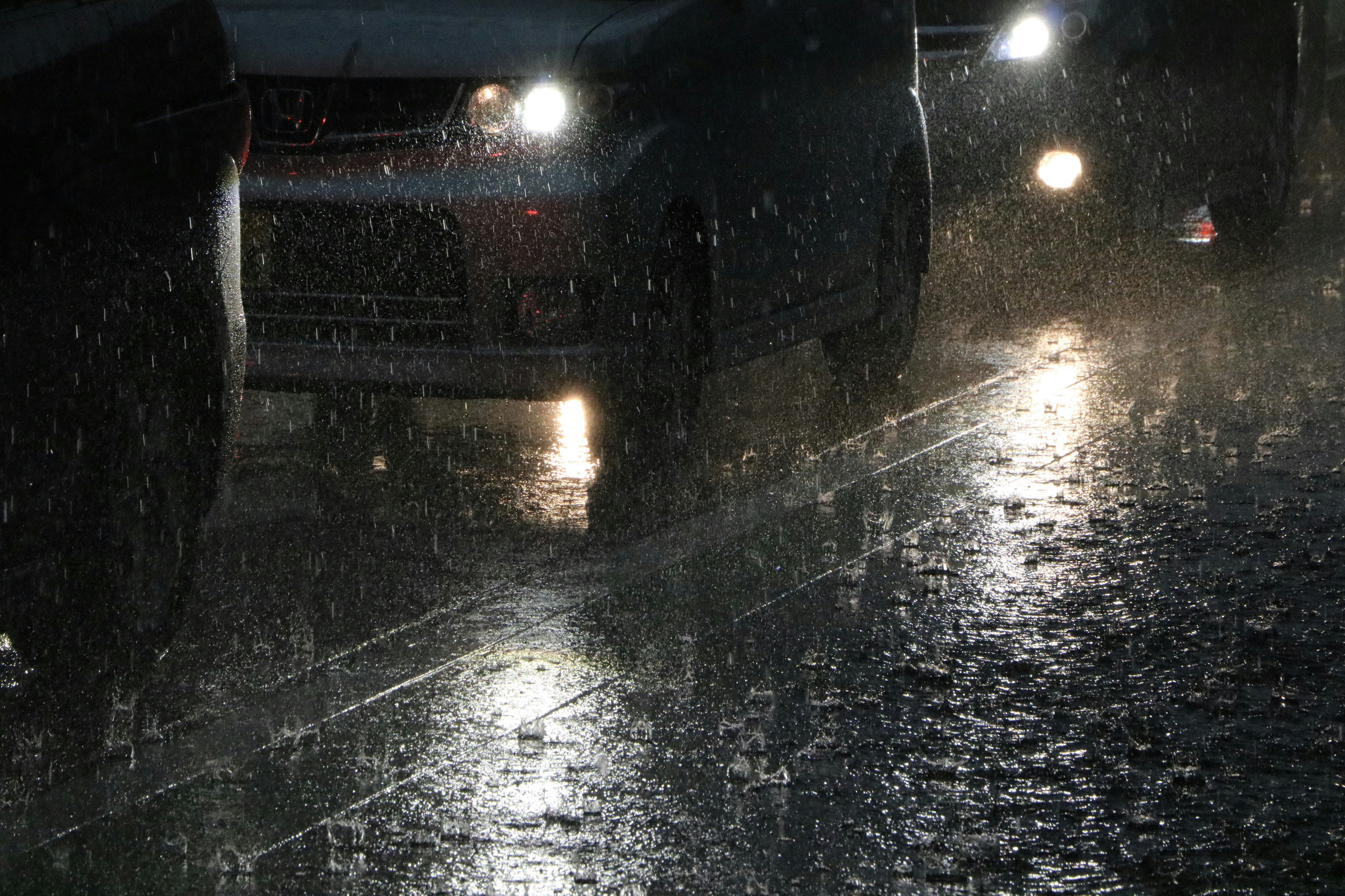 Cars with headlights on navigate a rain-soaked street, their lights reflecting on the wet surface.