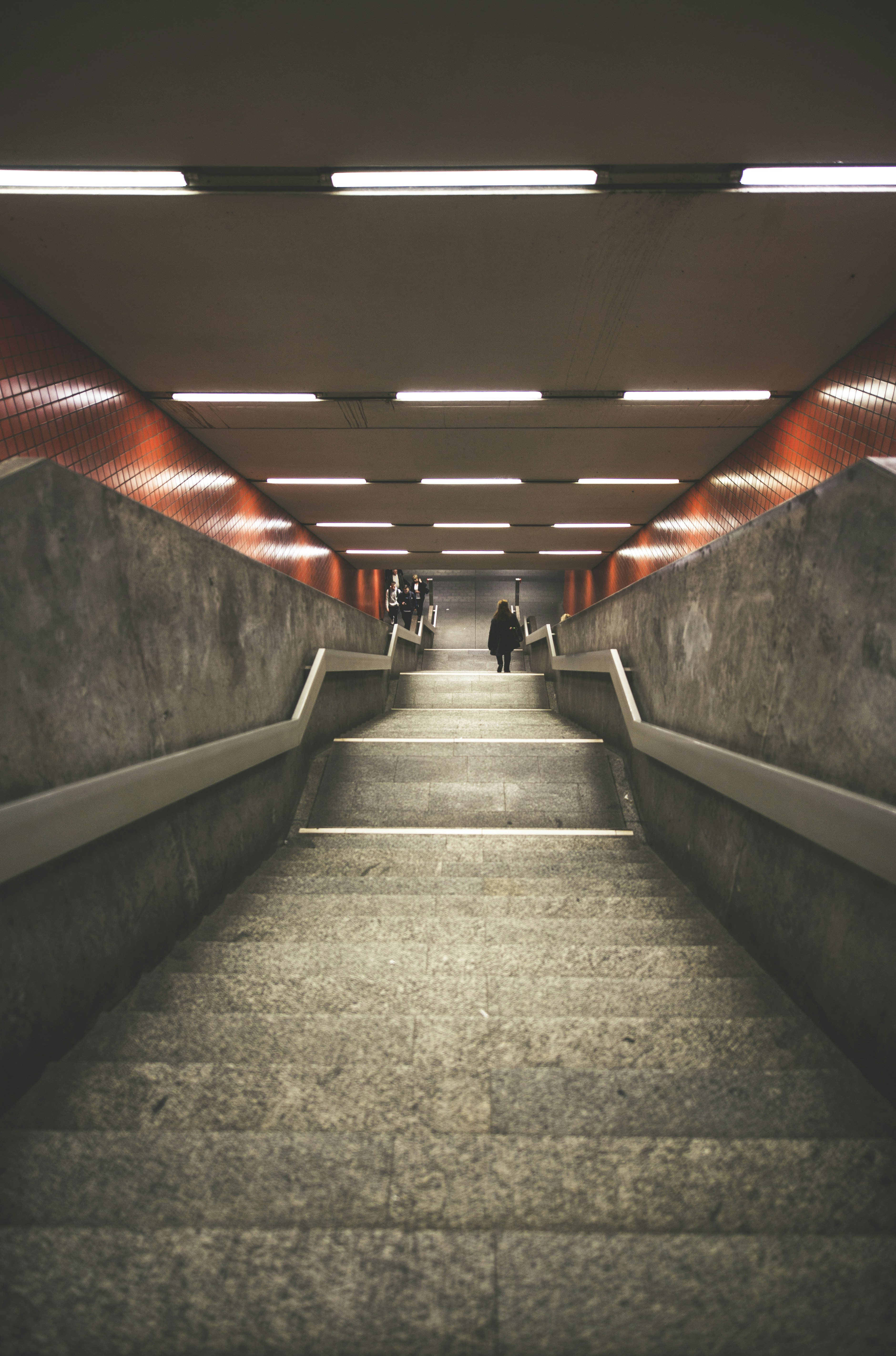 A solitary figure descends a wide staircase in a modern underground space, flanked by sleek concrete and vibrant red walls. Bright overhead lights illuminate the path ahead.
