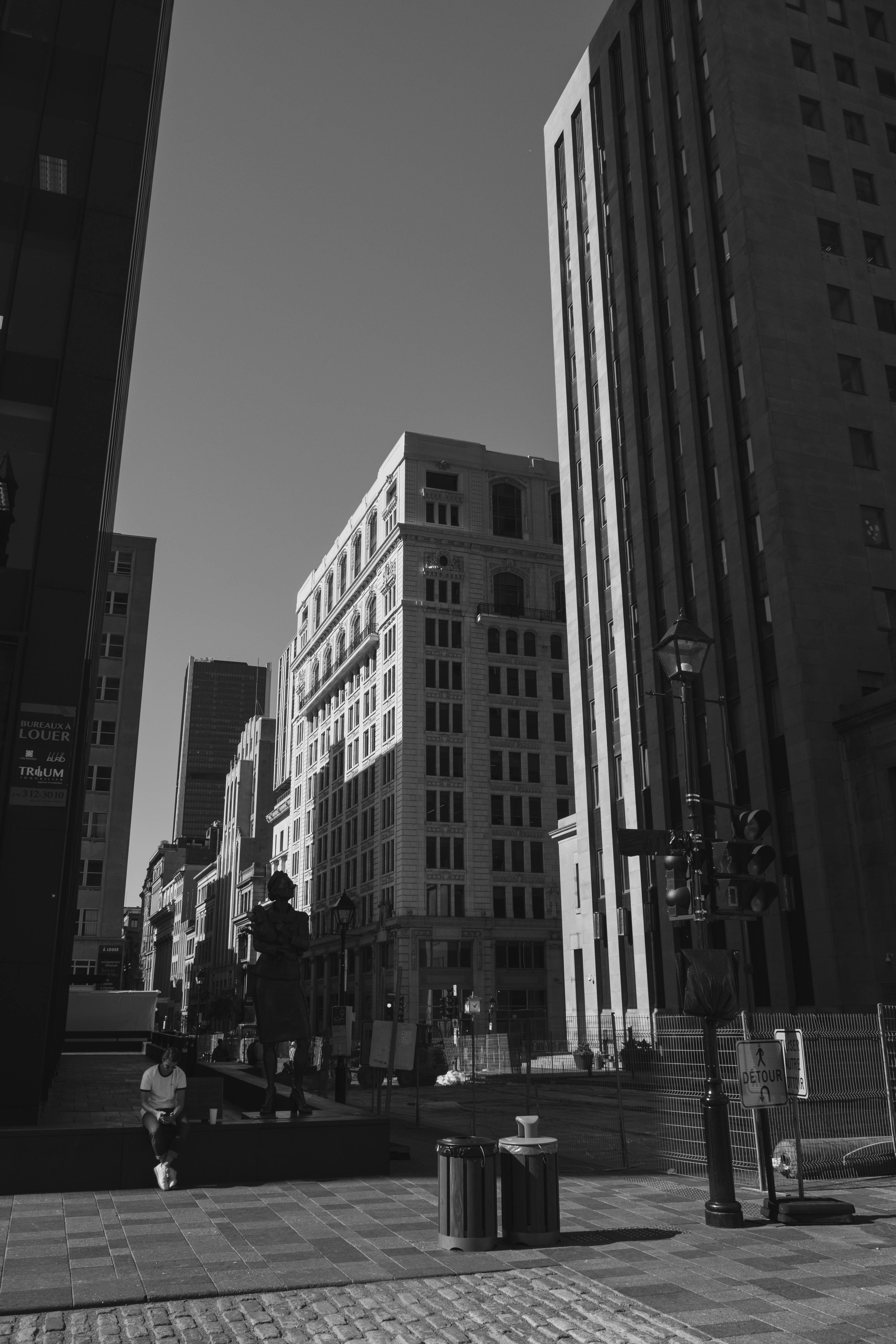 Monochrome cityscape showcasing towering buildings and a solitary figure seated on a bench. The interplay of light and shadows creates a dynamic urban atmosphere.