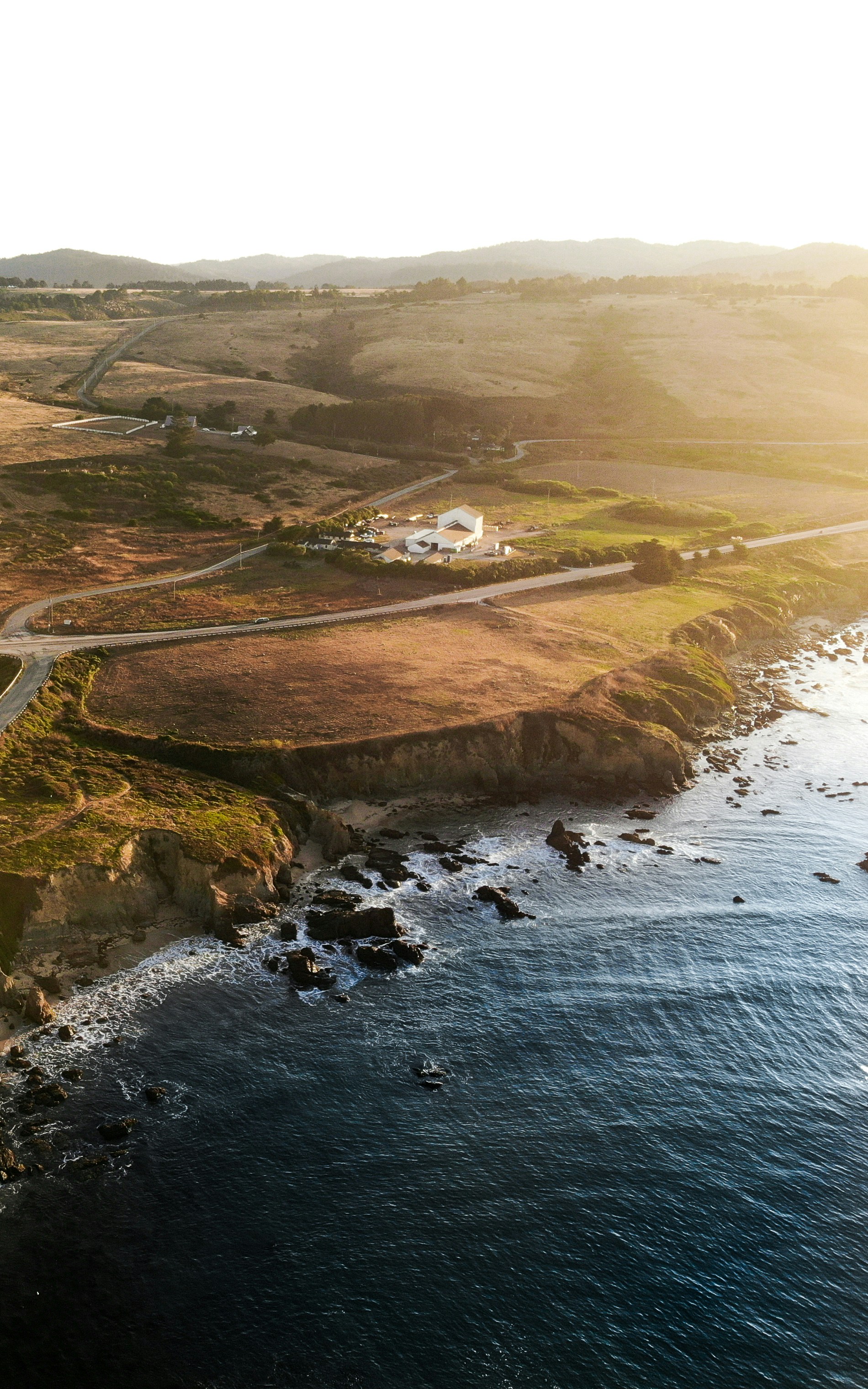 Aerial view of a coastal landscape featuring a house nestled against the rugged shoreline, with rolling hills in the background. The warm glow of sunset enhances the tranquil scene.