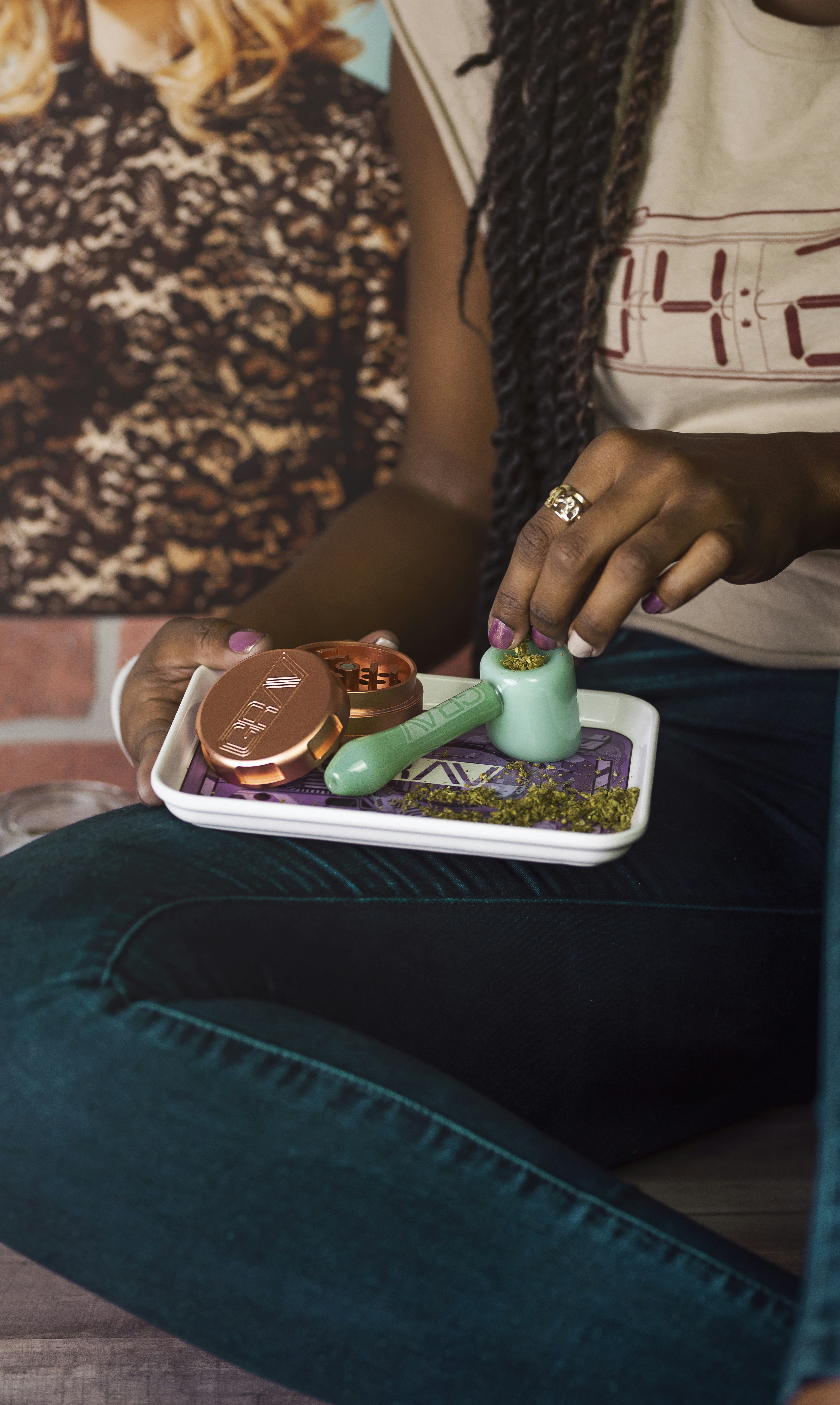 A hand delicately prepares herbs on a tray featuring a green pipe and copper grinder, set against a textured background.