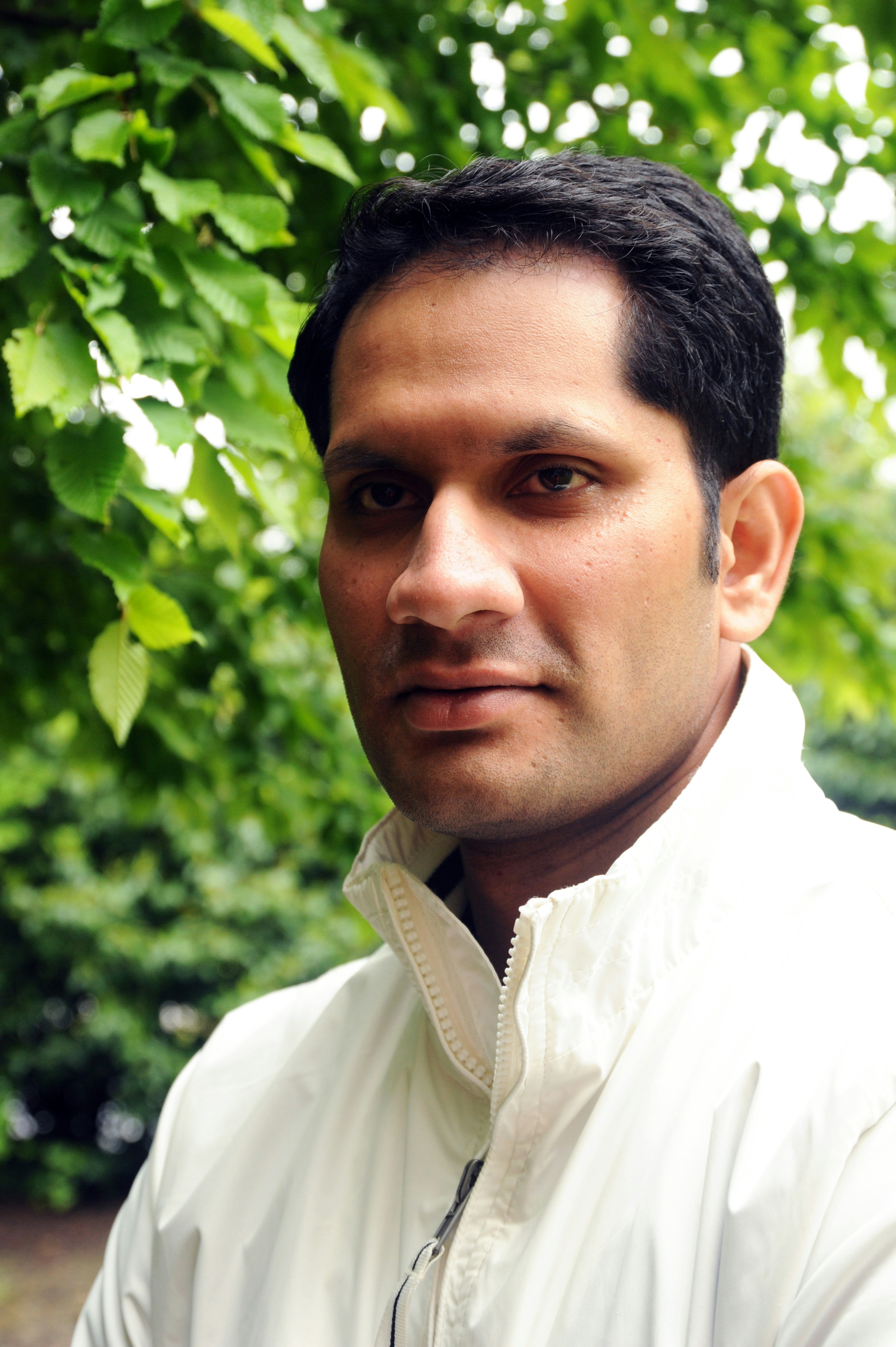 A man in a white jacket stands against a backdrop of lush green foliage, exuding a calm presence.