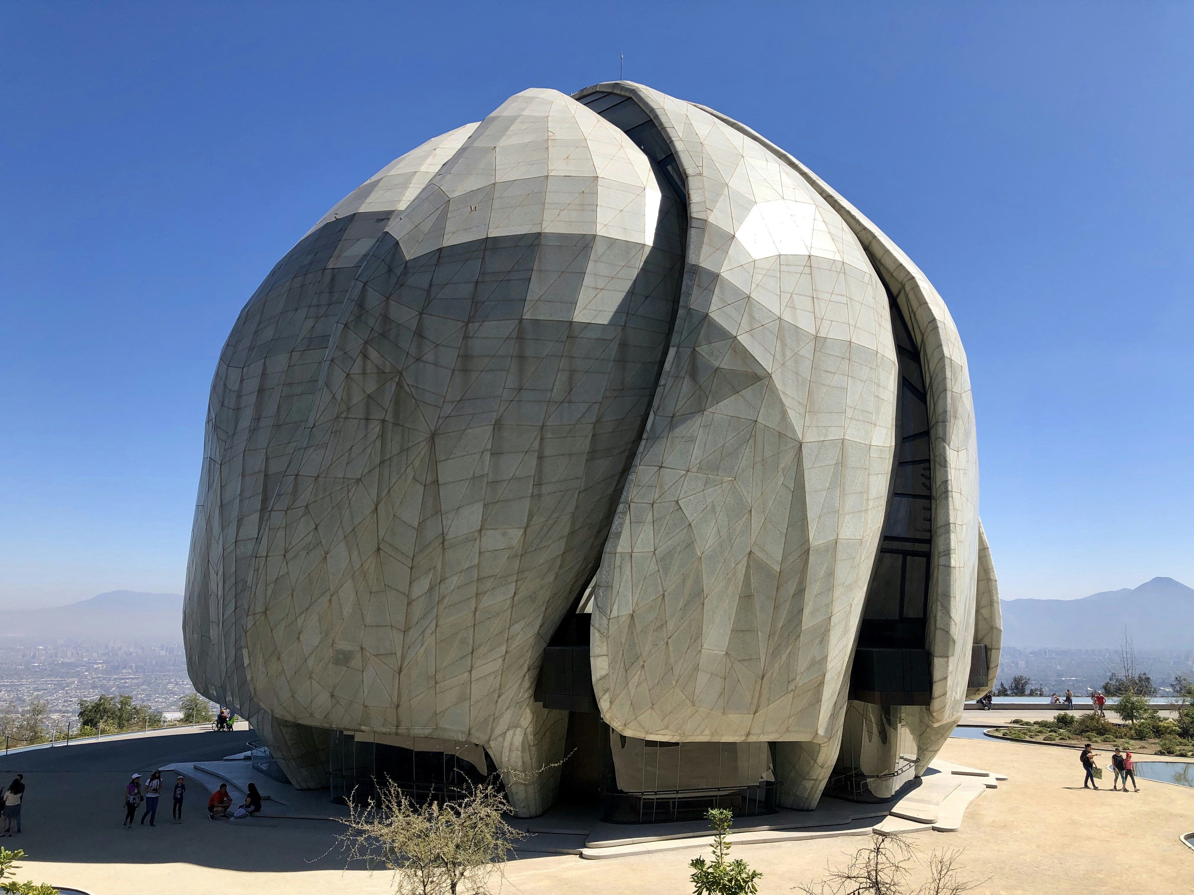 A striking architectural structure with a unique, organic shape set against a clear blue sky, showcasing innovative design elements. Visitors can be seen exploring the area around the building.