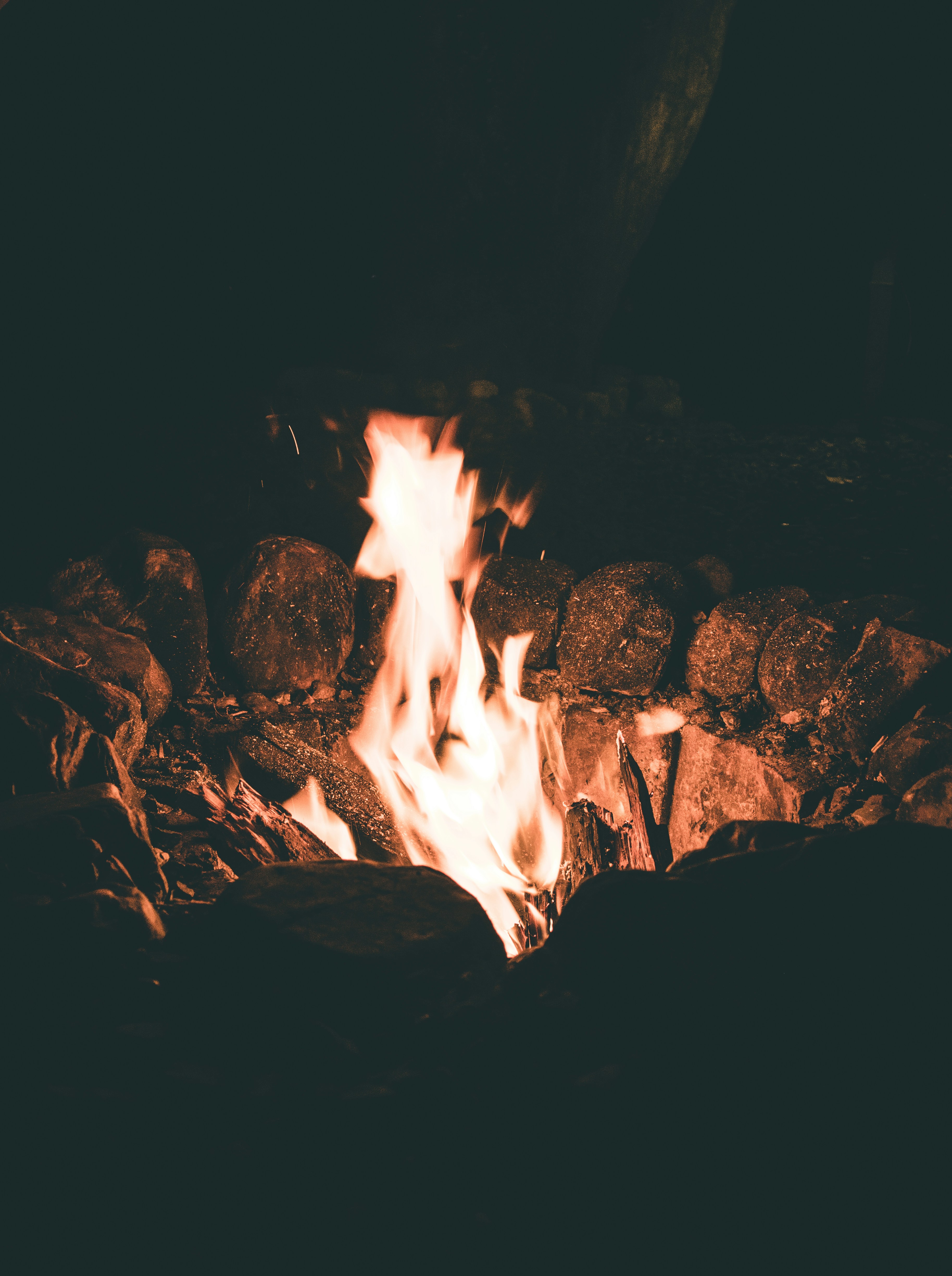 Flickering flames dance within a stone fire pit, surrounded by dark shadows of the night. The glow illuminates the rocks and sparks, creating a cozy atmosphere.
