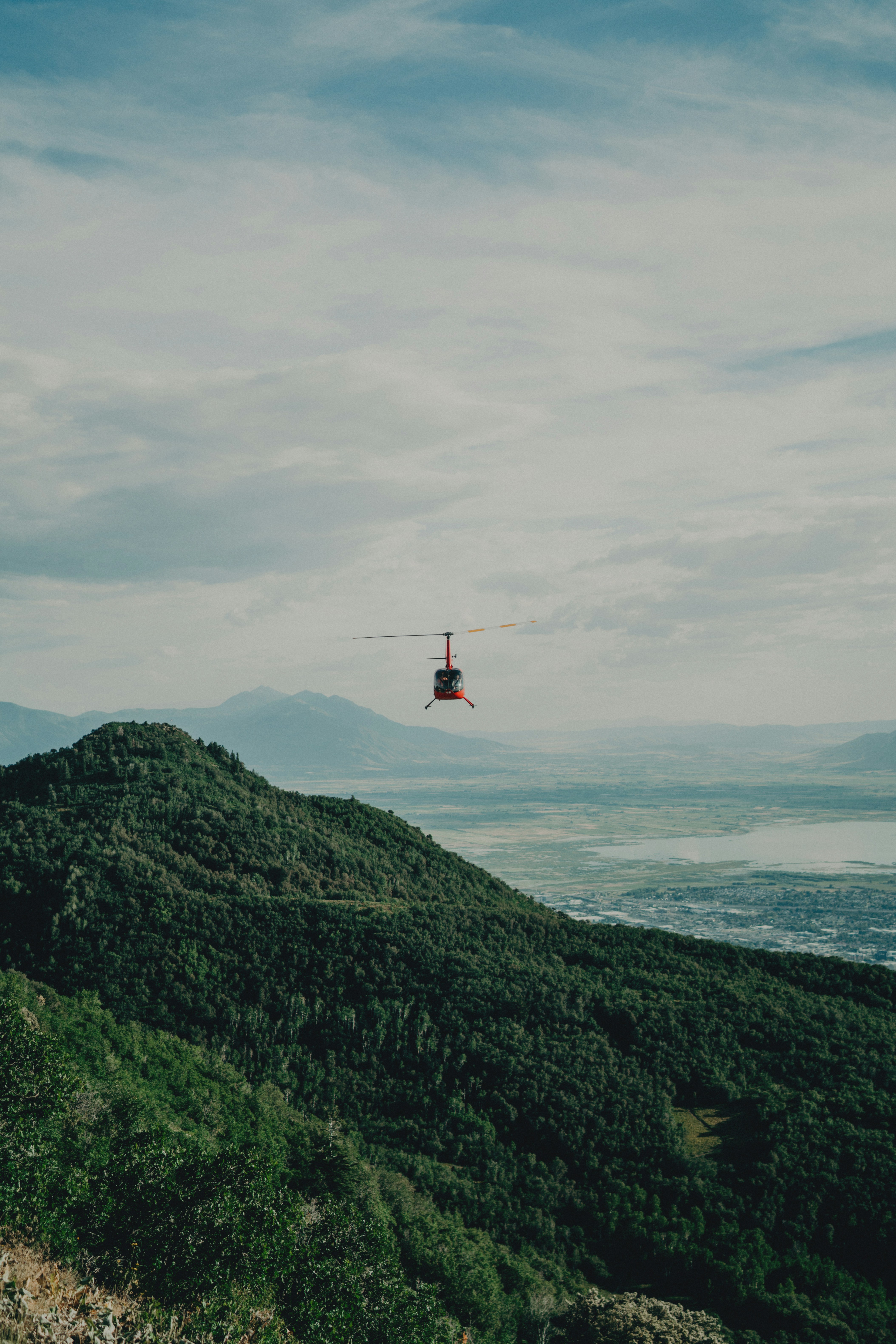 A helicopter hovers over a lush green mountain landscape, showcasing the vastness of nature below. The scene captures the harmony between technology and the wilderness.