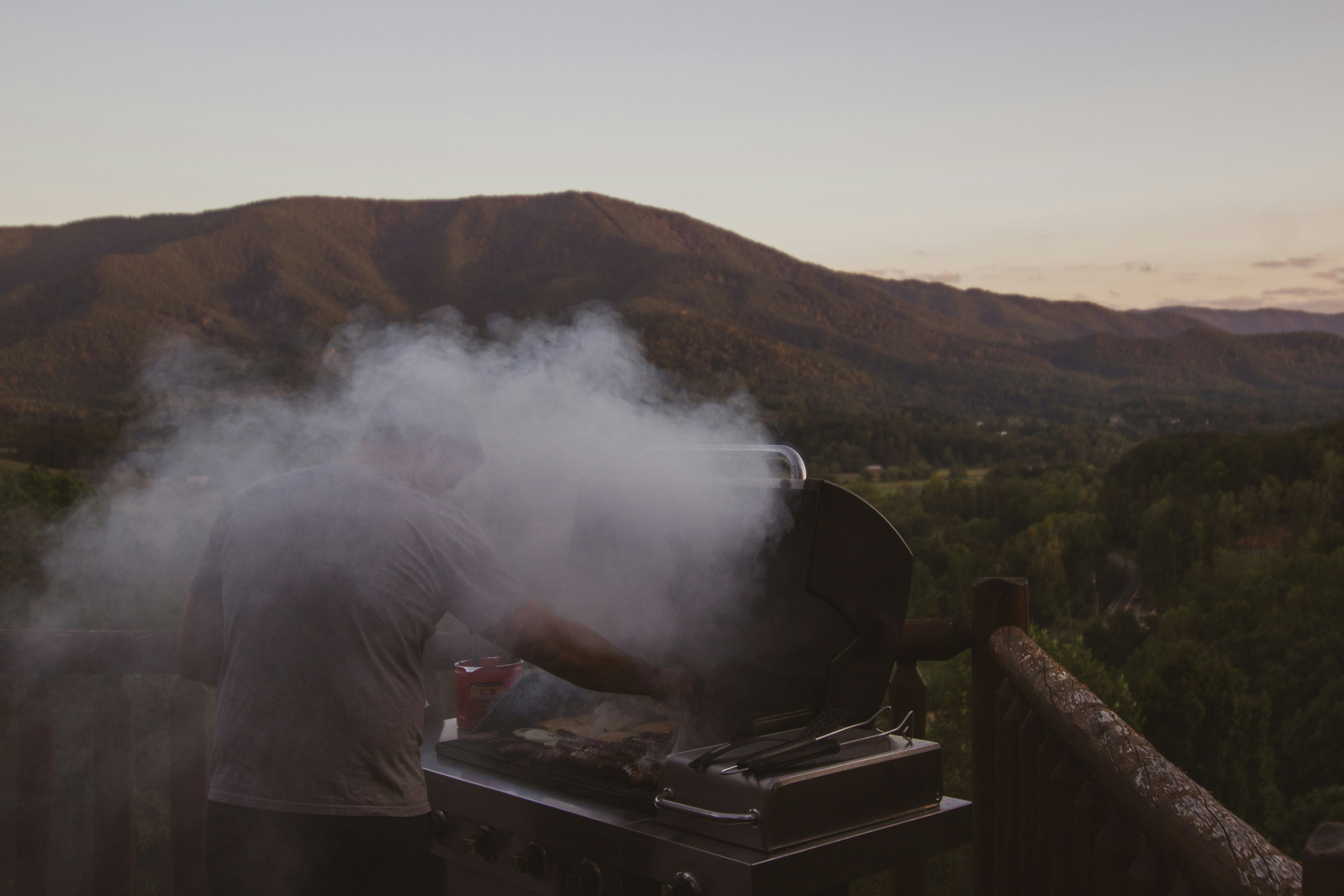 A person tends to a grill, surrounded by a smoky haze and a panoramic mountain view during twilight.