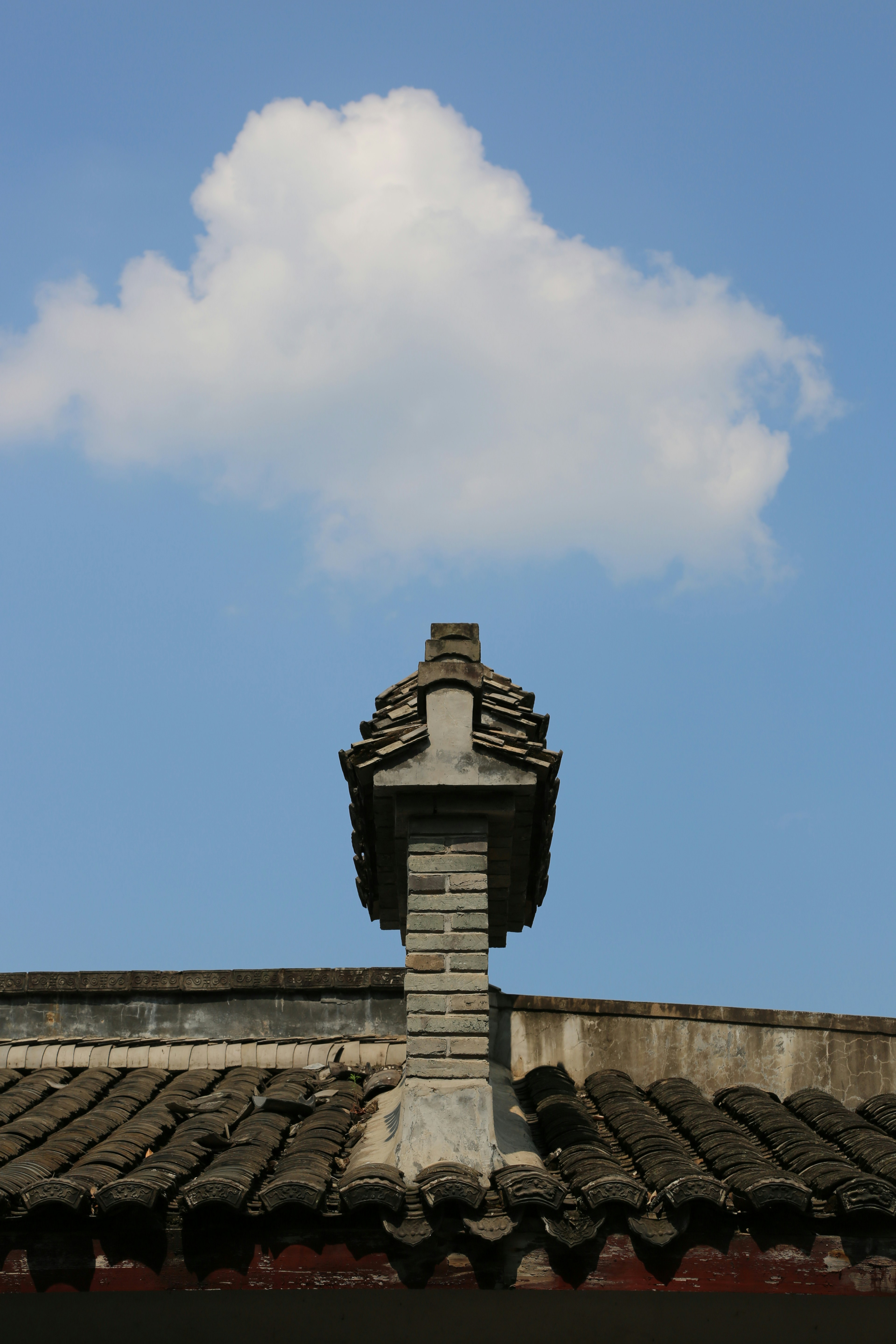 Traditional architectural chimney rising from a tiled roof under a clear blue sky with a fluffy cloud above. 