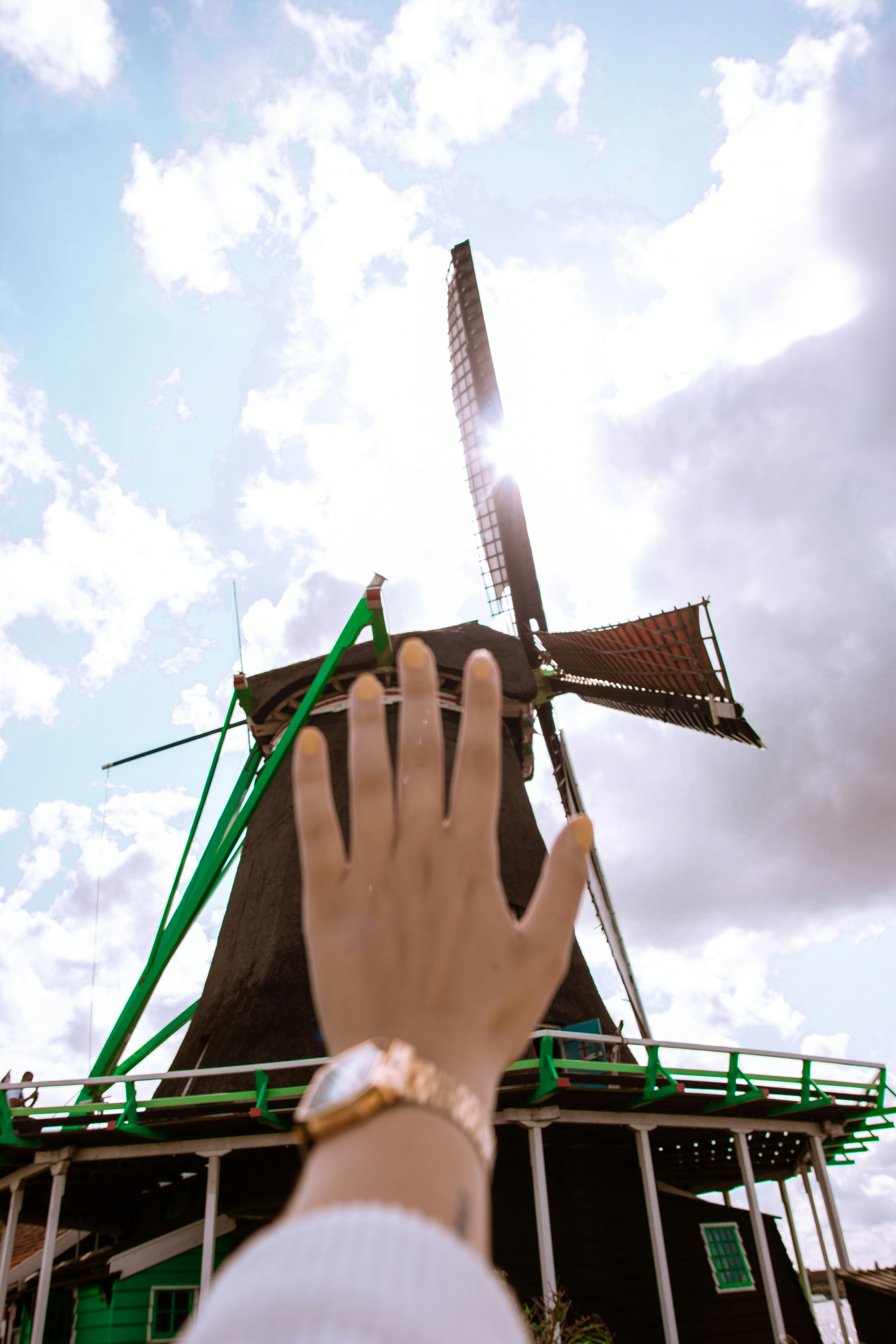 A hand reaches out towards a traditional windmill, framed by a bright sky and dynamic clouds. The windmill's blades catch the sunlight, creating a striking silhouette.