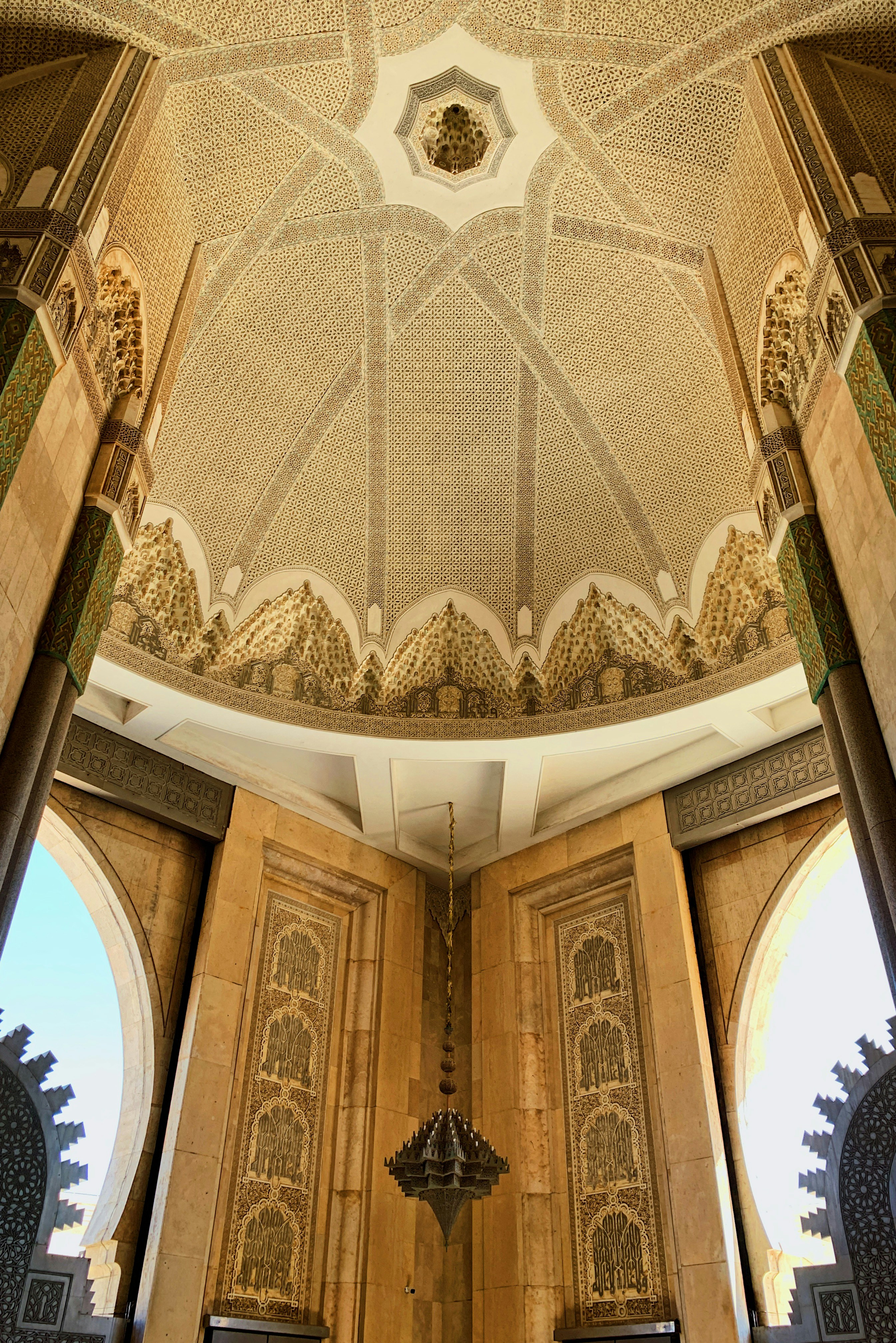 Intricate ceiling design adorned with geometric patterns and a central chandelier, framed by grand arches. The craftsmanship showcases traditional artistry.